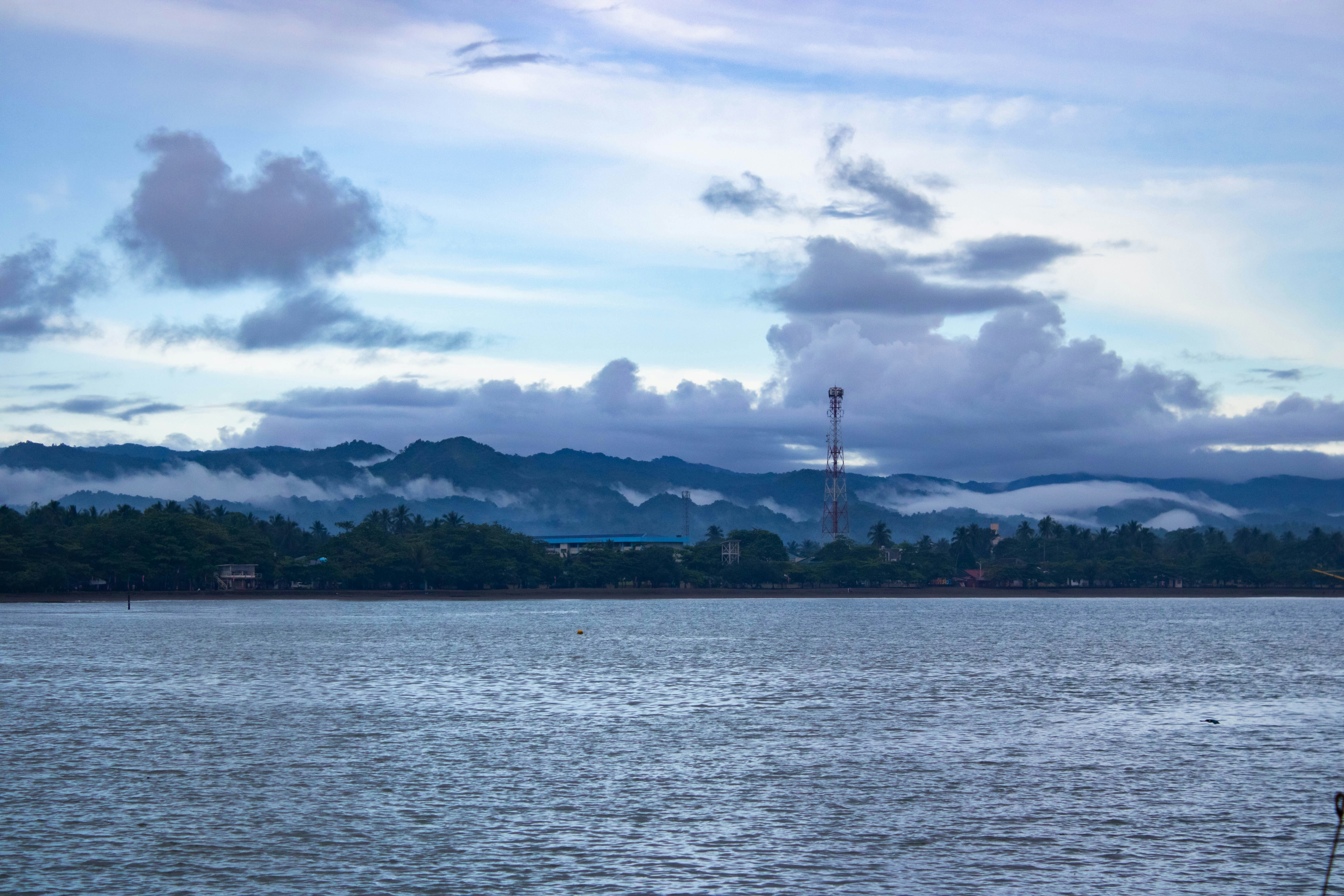 Coastal scene with calm water, distant shoreline, low-lying mist, and a tall structure on the horizon; a serene landscape emphasizing atmosphere. Logo-like skyline appears natural and unaltered.