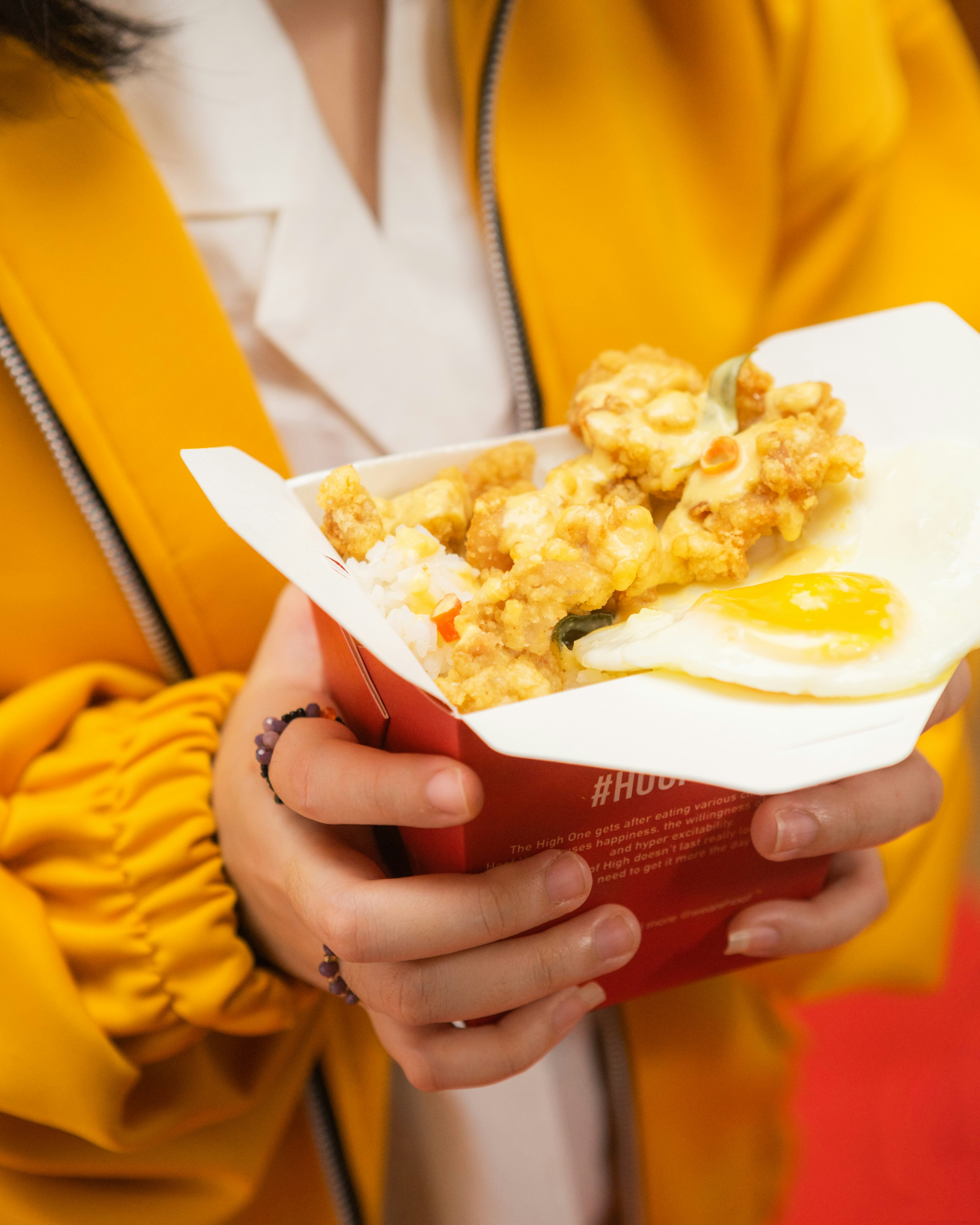 Crispy fried chicken bites with a sunny-side egg in a red takeout box, held by a person in a vibrant yellow jacket.