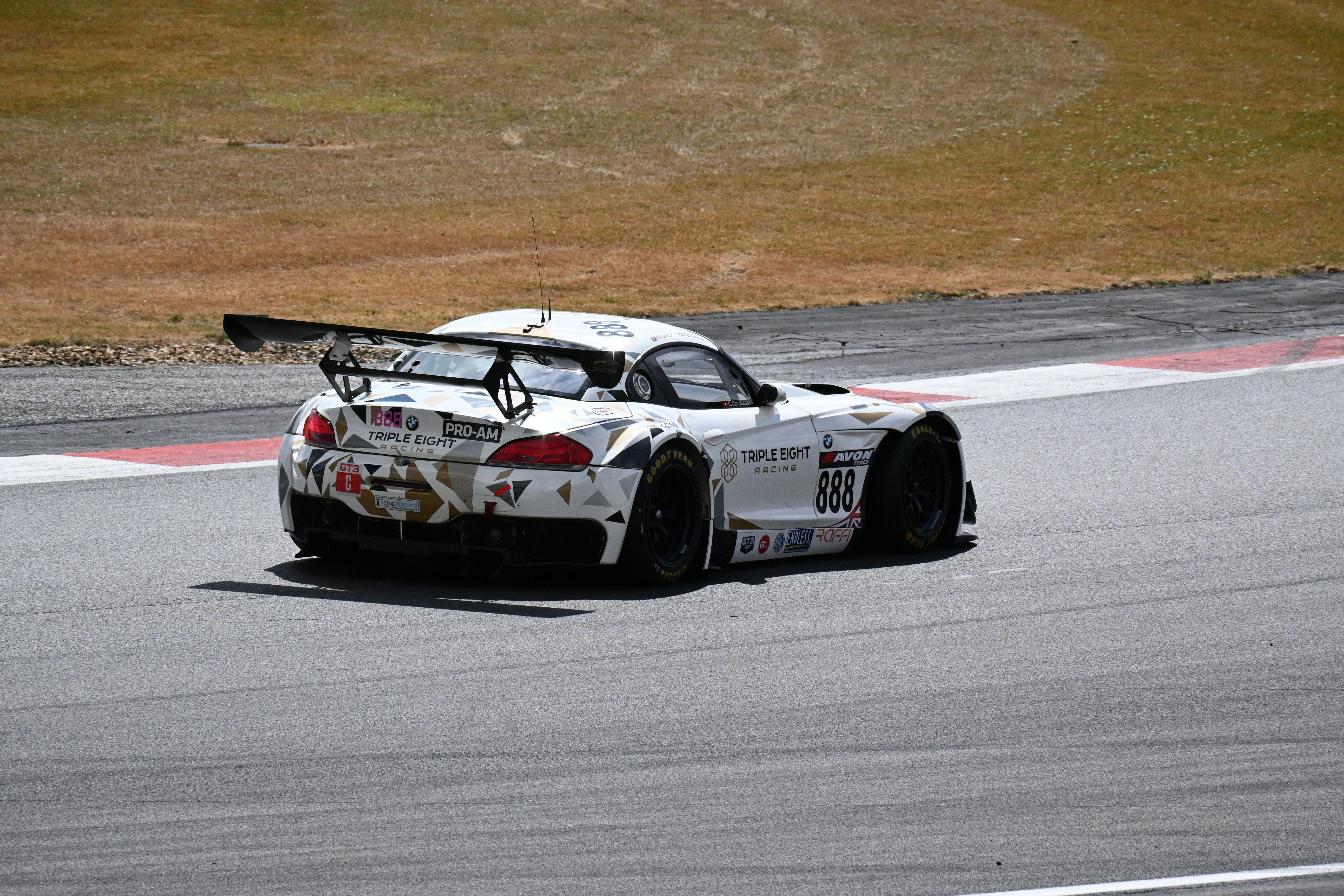Race car with livery braking through a sunlit track; the image centers on a low-slung GT car amid asphalt and grassy runoff.
