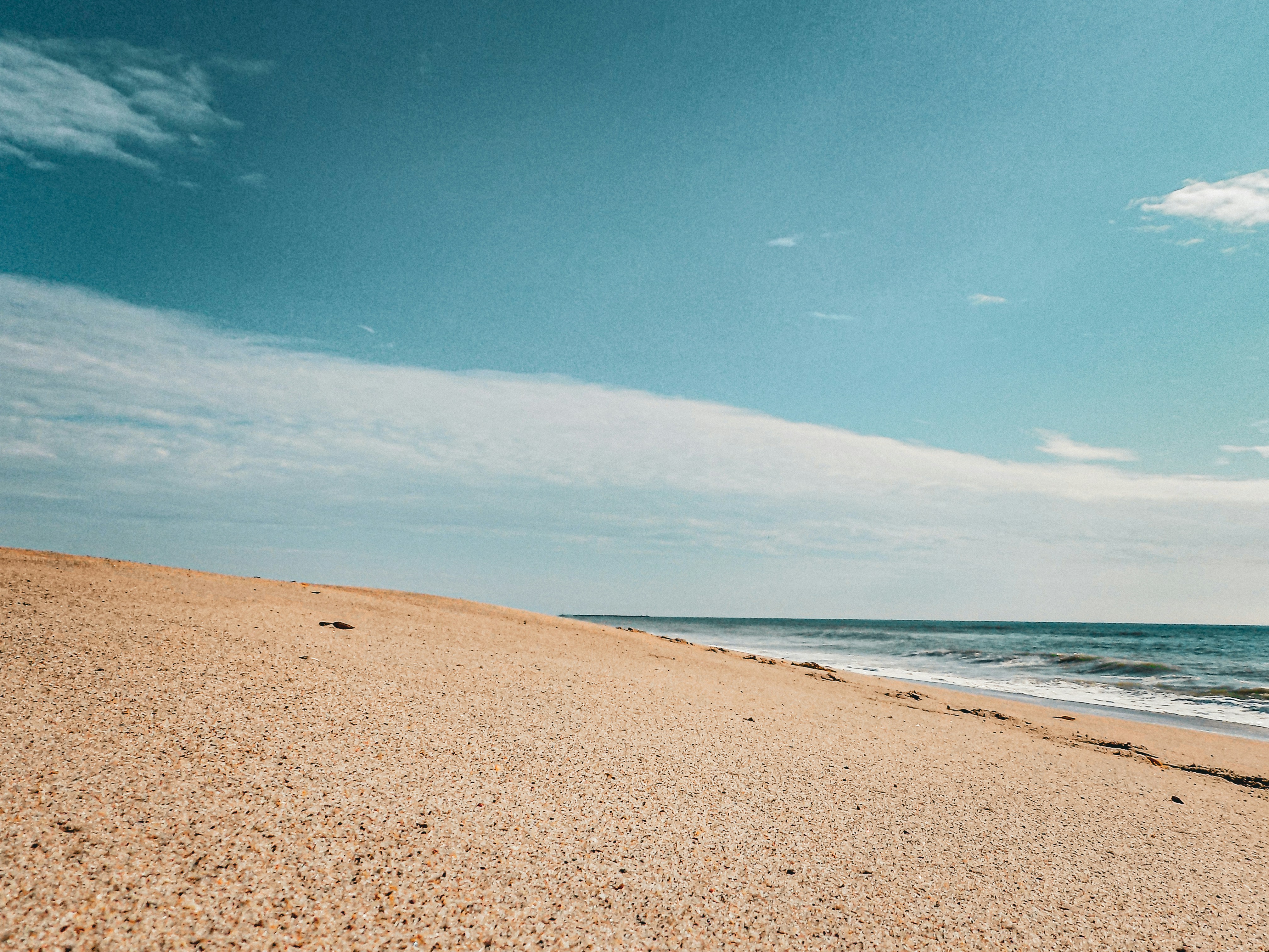 Sunlit sandy shoreline meets calm sea with a clear blue sky; a serene beach scene devoid of people, highlighting expansive horizon and gentle waves. Photograph appears as a natural landscape shot.