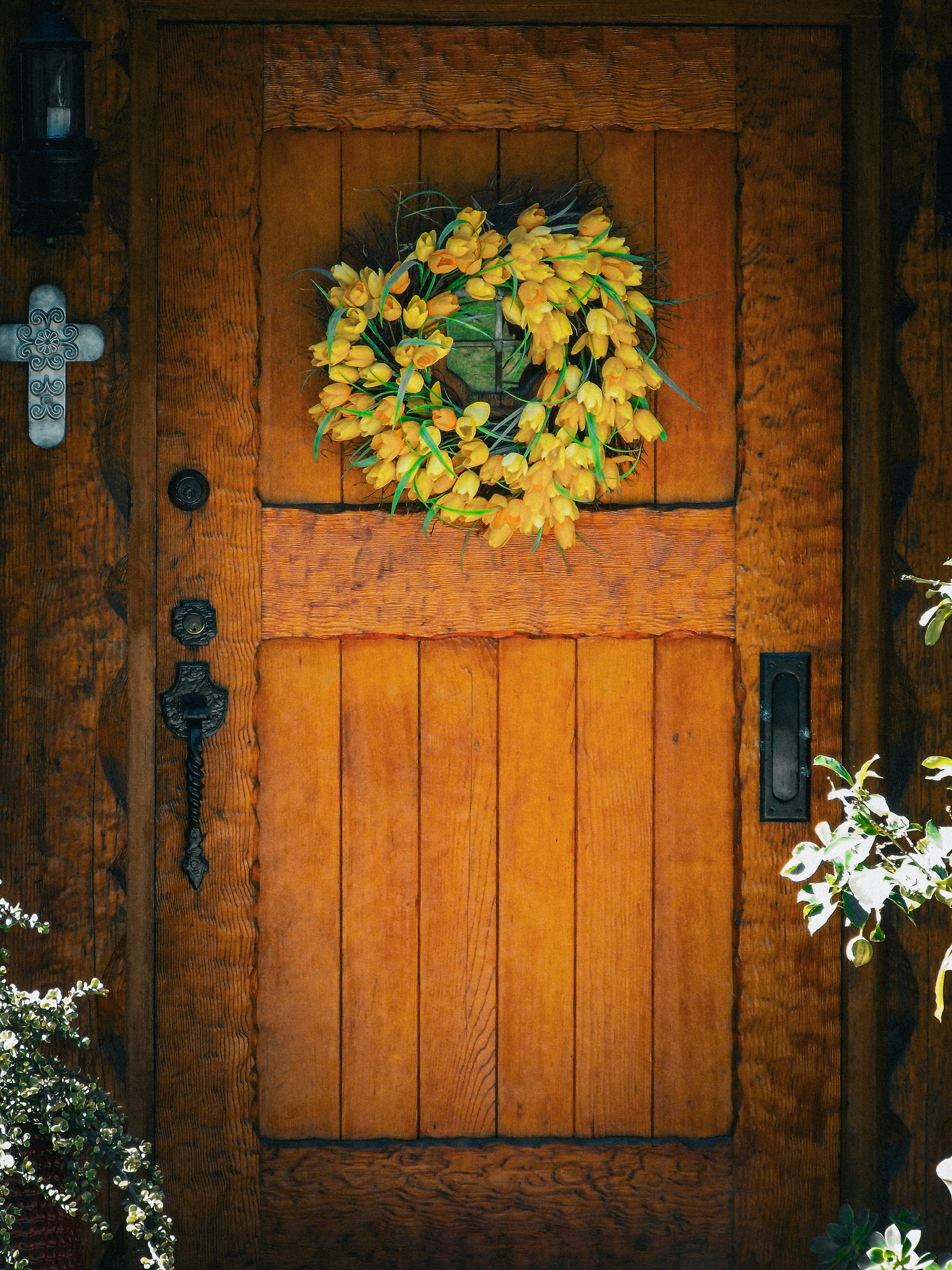 A warm-toned wooden door features a vibrant yellow wreath in a square window, framed by wrought-iron hardware and surrounding greenery.