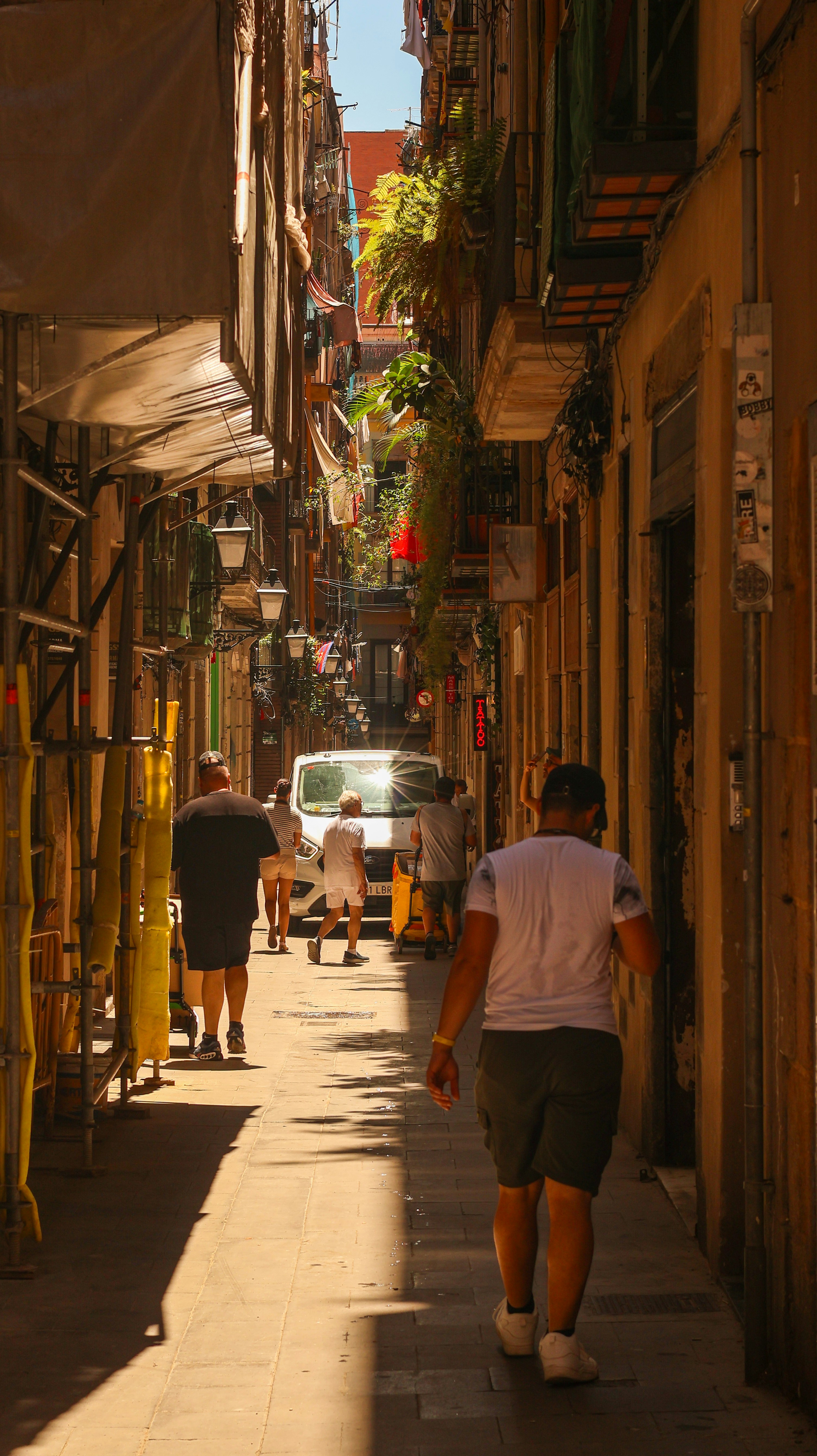 Sunlit alley with pedestrians strolling along a narrow street flanked by old buildings; a bright van in distance and hanging plants add color and depth. Urban scene photograph, capturing daily activity in a historic lane.