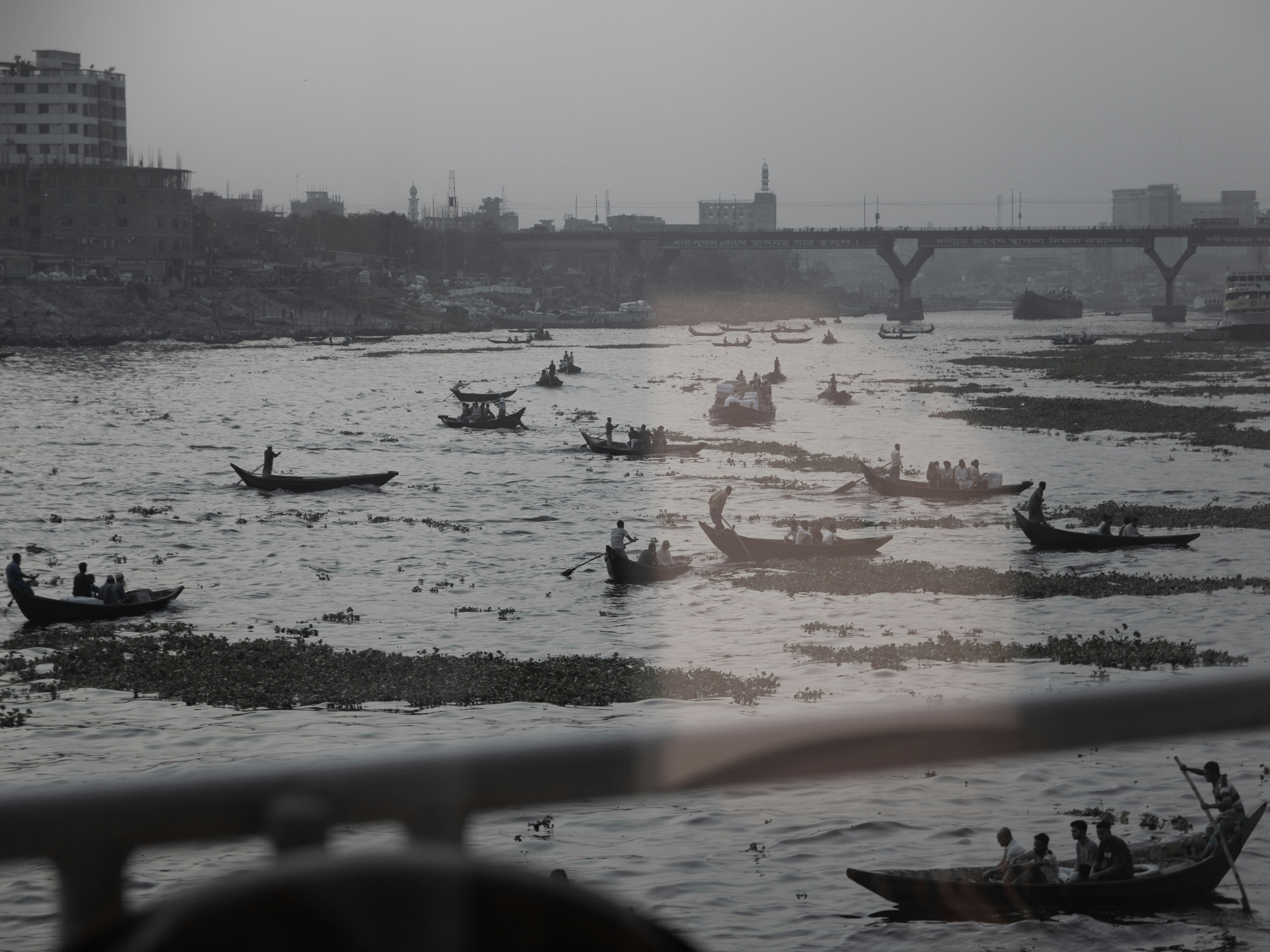A hazy, sepia-toned wide shot captures a busy river crossing dotted with dozens of small wooden boats, each carrying passengers between the shores. A city skyline with a bridge, minarets, and rooftop buildings lines the horizon under a soft, overcast sky. Thick patches of floating water hyacinth spread across the surface, adding texture to the muted, golden light of what appears to be early morning or late afternoon — a vivid slice of urban river life, likely along the Buriganga River in Dhaka.