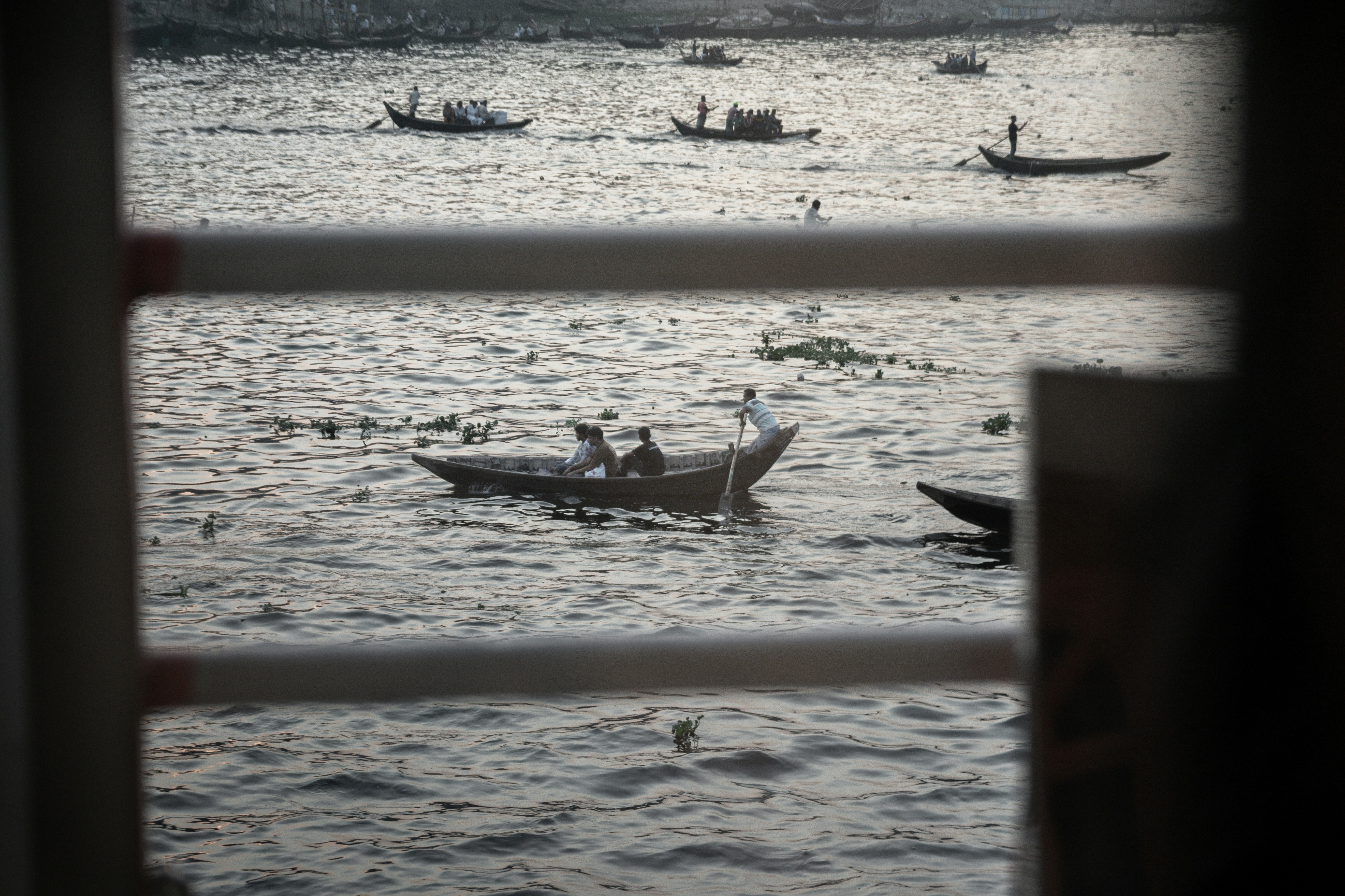 Small wooden boats ferry passengers across a busy river, shot through the wooden slats of a window or railing. The water glimmers with hazy, backlit light — likely early morning or late afternoon — while a boatman poles a crowded canoe-style vessel in the foreground and several similar boats dot the background near what looks like a busy waterfront or dock area. The composition, with the frame's slats cutting across the shot, gives it a candid, voyeuristic feel — like a quiet observed moment of daily river life, possibly in South Asia (the boat style is reminiscent of the Buriganga River in Dh