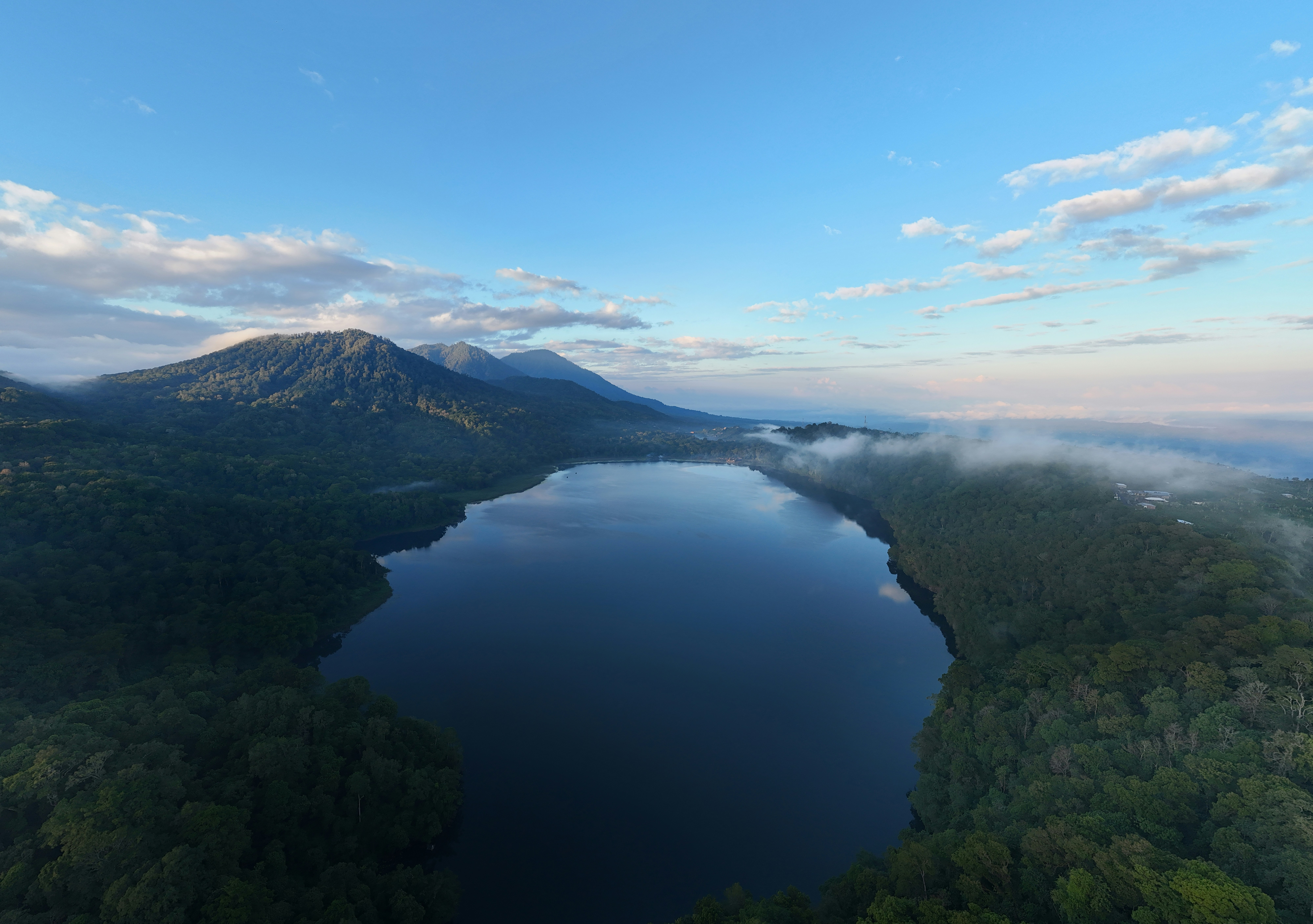 Open-water lake carved between forested ridges under a clear sky, with distant mountains; drone capture highlighting the reflective water and surrounding greenery. (Aerial shot, drone landscape).