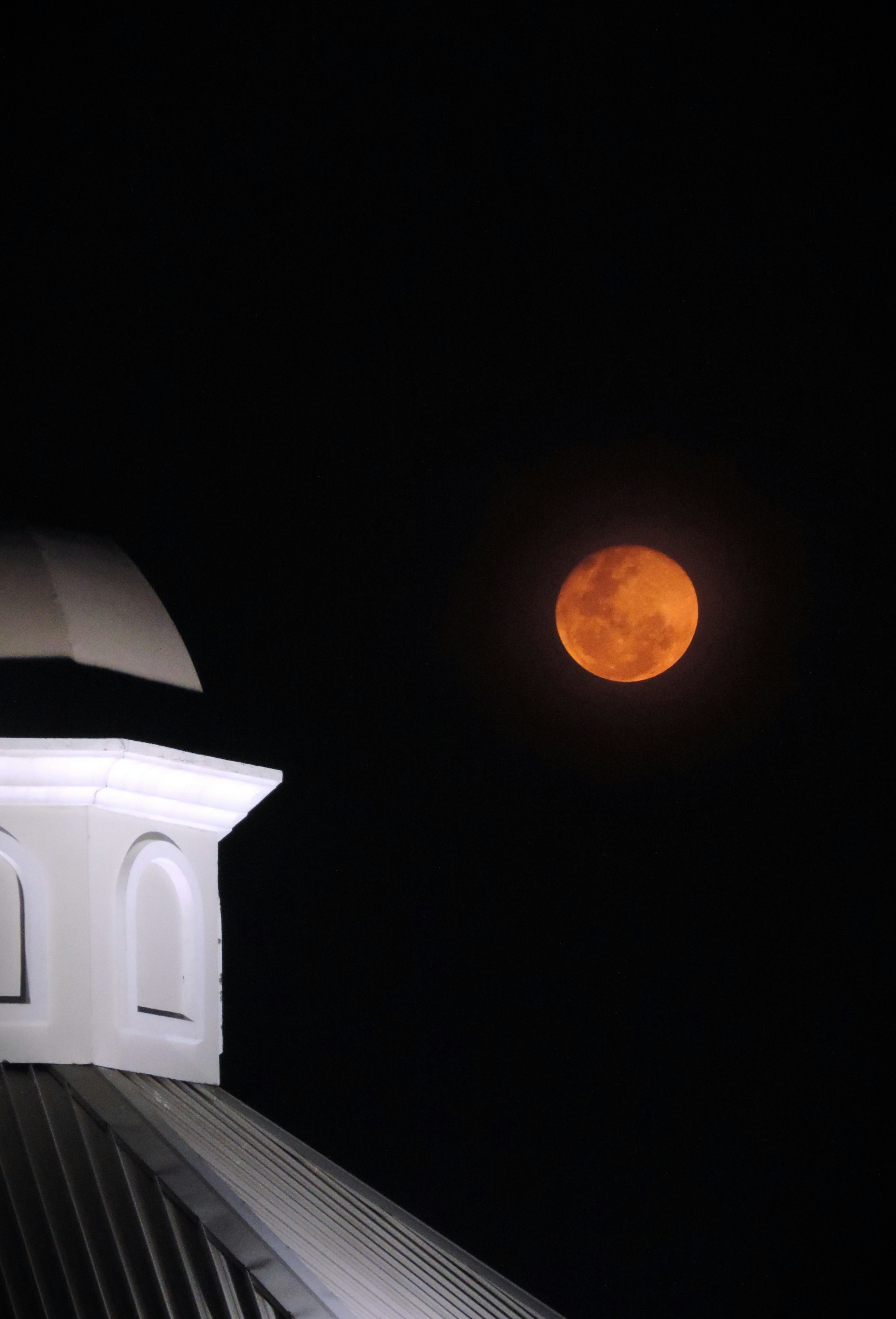 A copper-toned lunar glow lingers in the night sky beside the white architectural roof, captured with a telephoto reach. The scene emphasizes the contrasting structures against darkness.