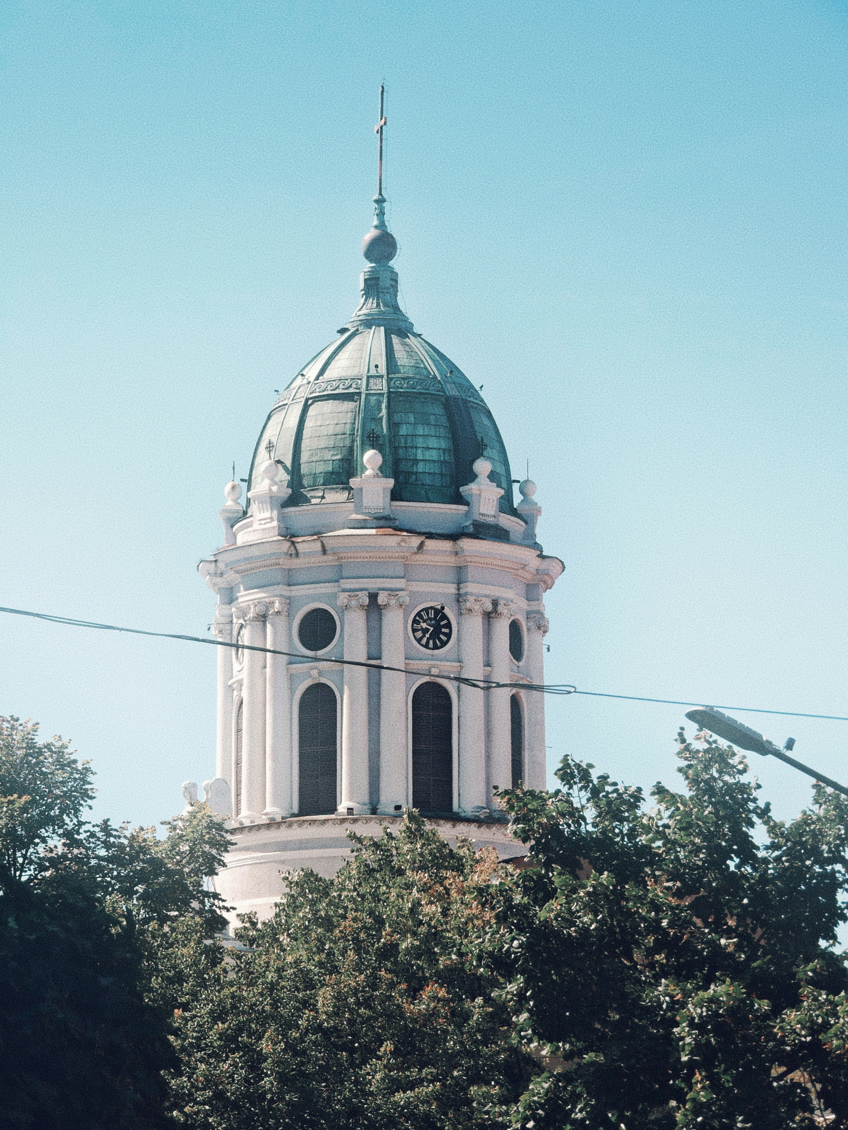Towering clock tower with a green copper dome rises above leafy silhouettes against a pale blue sky; a historic municipal building framed by cables and urban foliage.