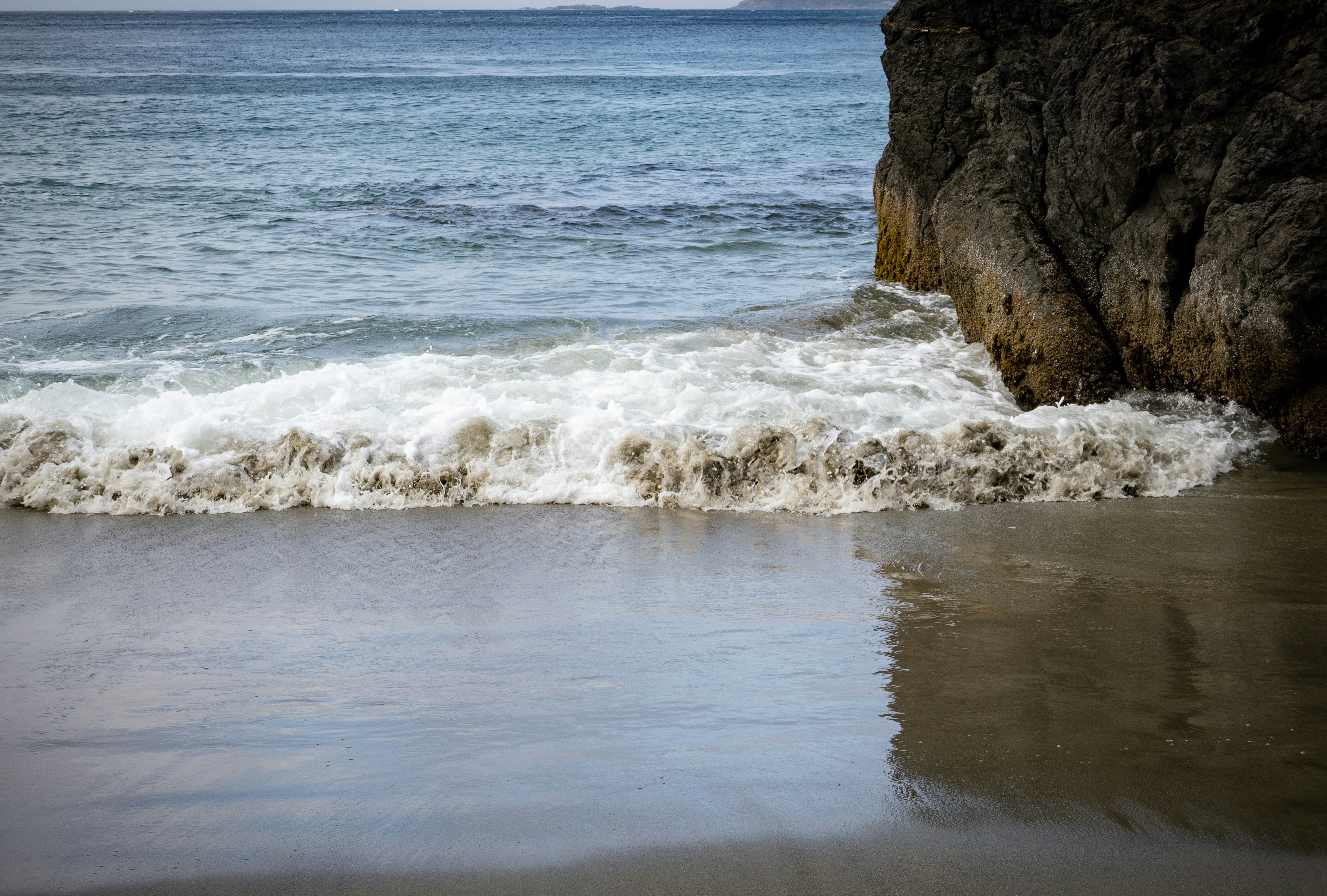 Coastal shoreline with waves breaking along a sandy beach and a prominent rocky outcrop on the right.