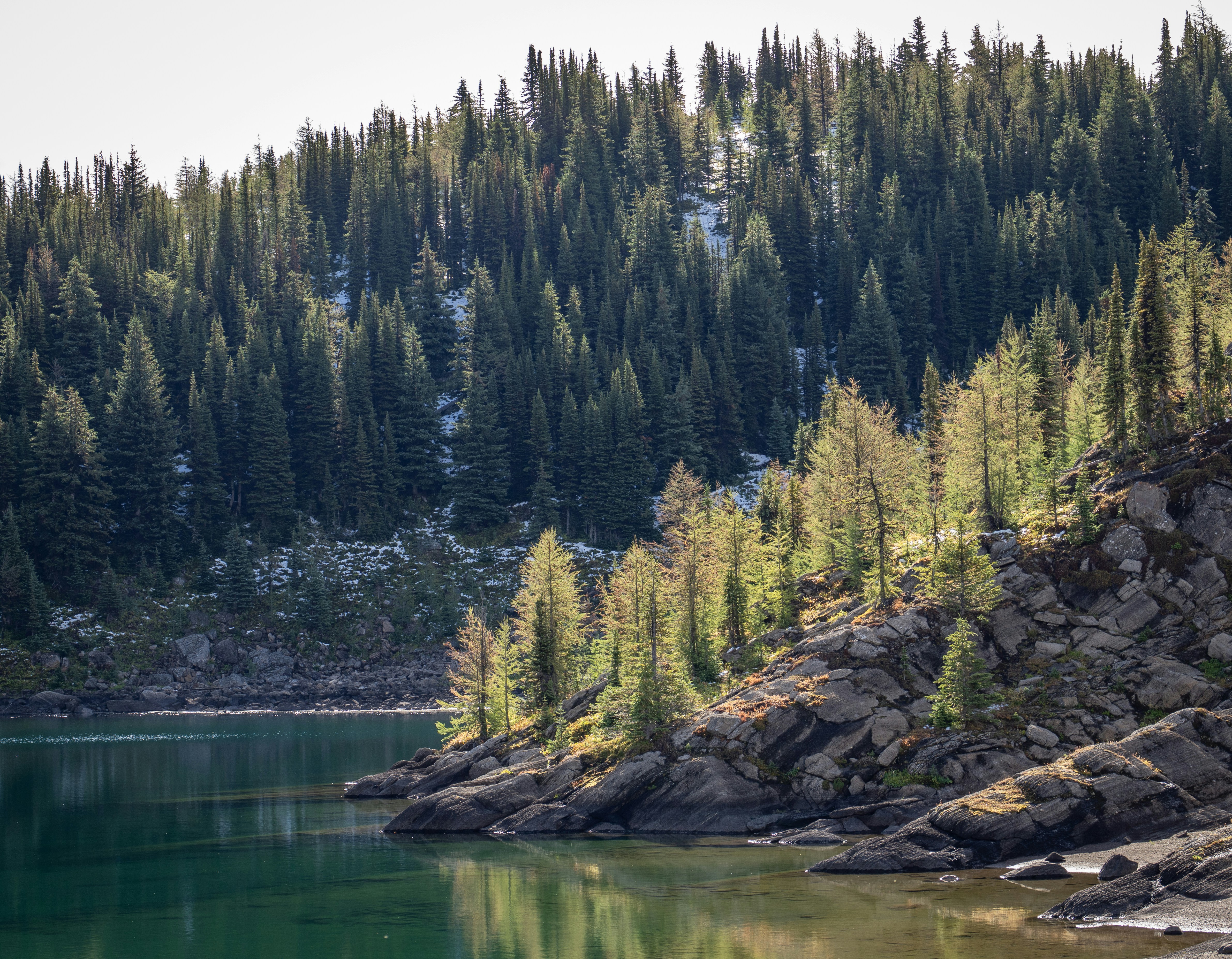 Glacial lake with emerald water and sunlit pine trees on rocky shoreline, framed by forested hills.