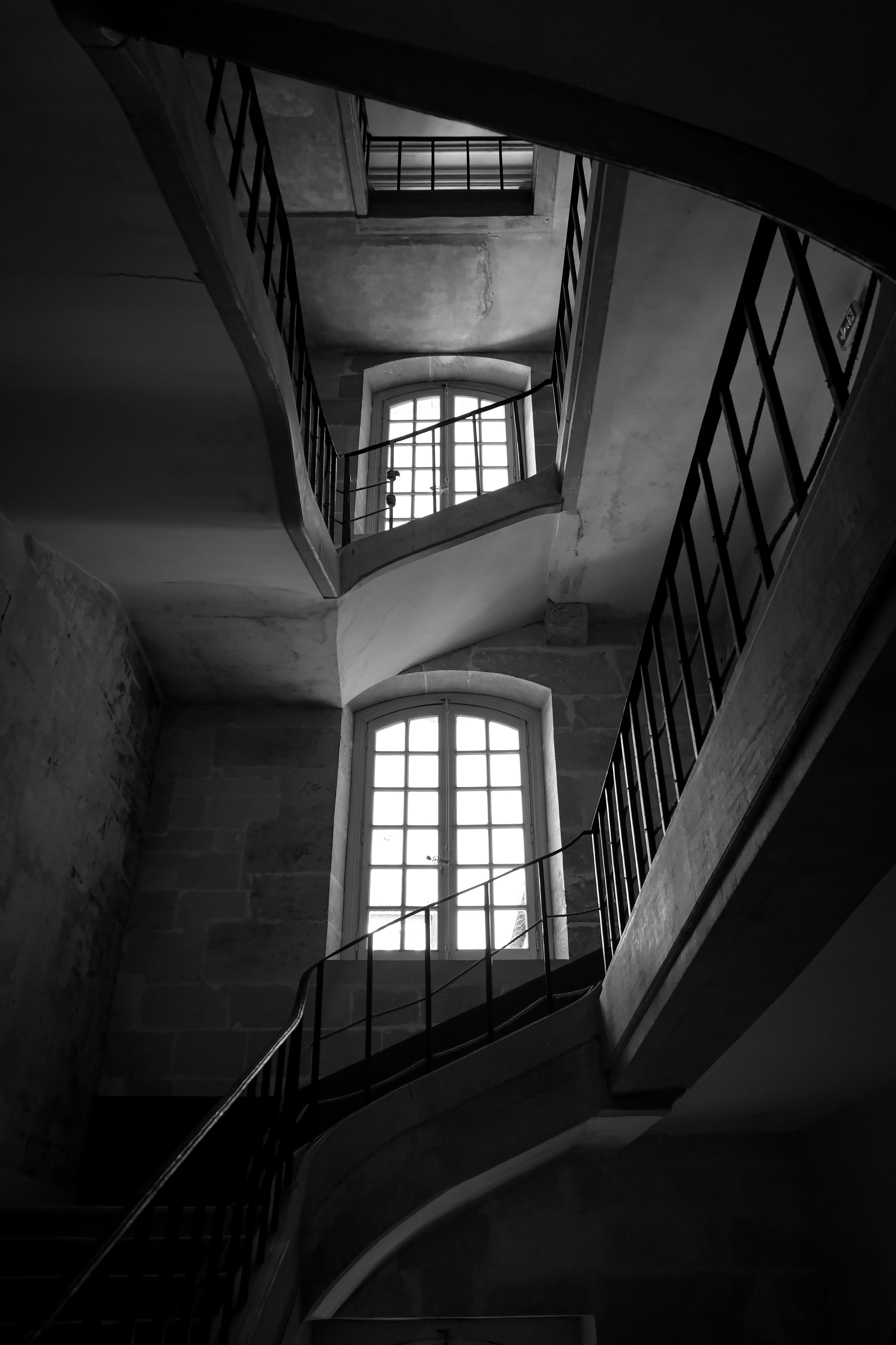 A black‑and‑white architectural interior capturing the curved lines of a staircase and the sharp geometry of large window panes. Strong contrasts between light and shadow highlight the structure’s rhythm, creating a quiet, cinematic moment shaped by form, depth, and minimalism.