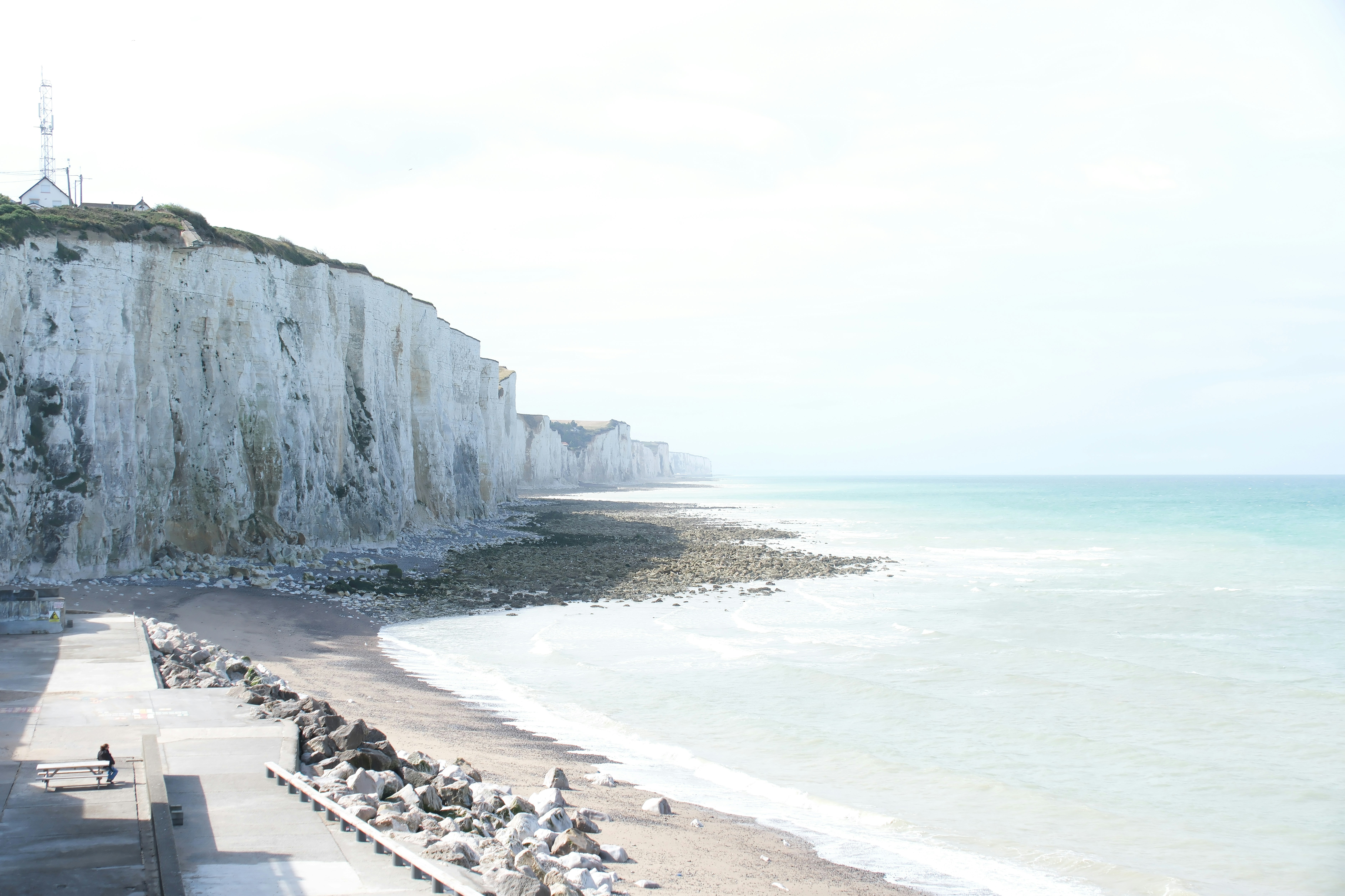Cliffs line a calm shoreline with a concrete promenade and a rocky jetty meeting turquoise water at the horizon, captured in soft daylight. A quiet coastal scene with a lone figure near the steps.