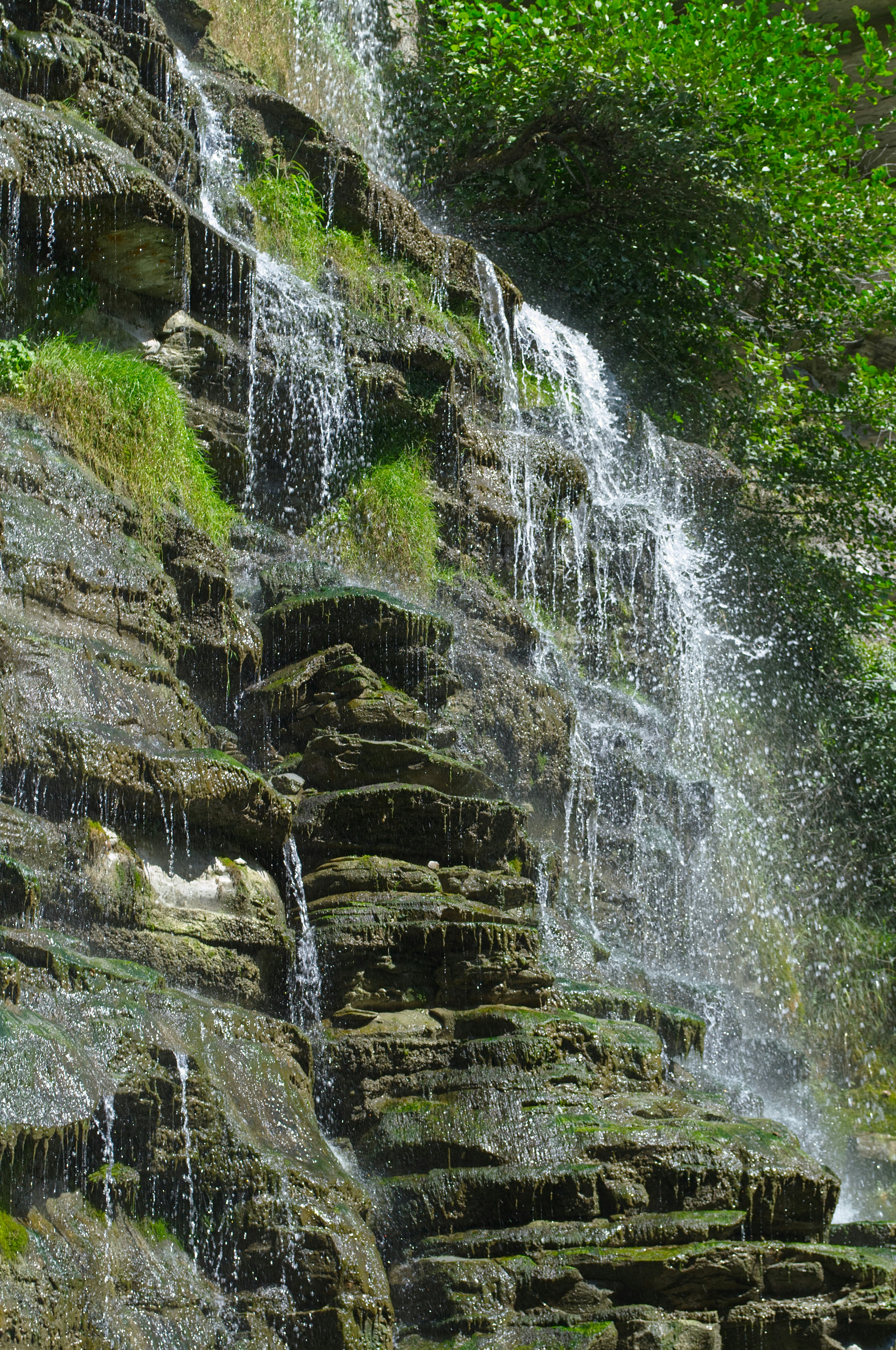 Mossy layered rock stairs catch a delicate spray of waterfall in a lush, green setting. A natural scene capturing the force and texture of cascading water across stone.