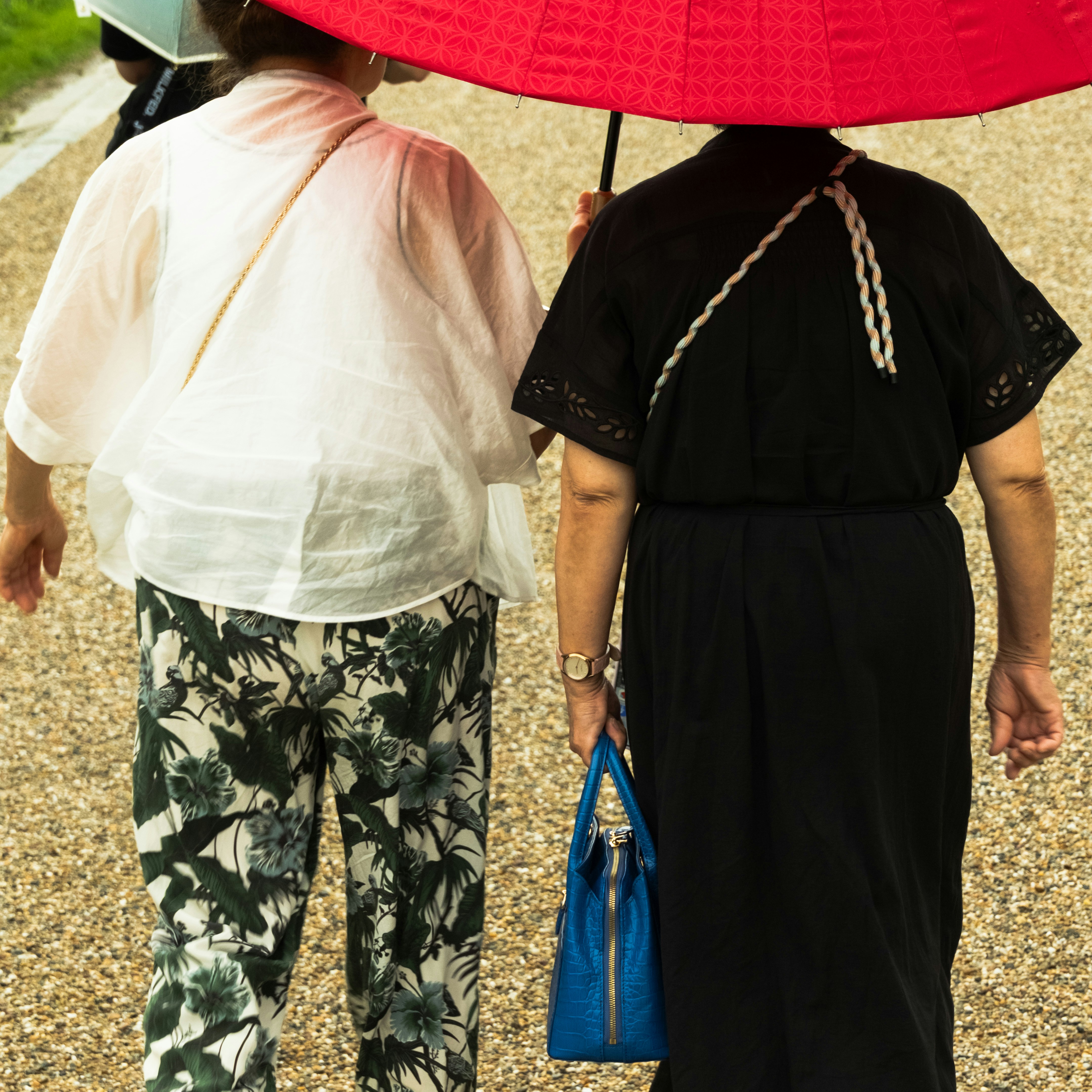 Two women walk away under a red umbrella on a gravel path, one in a white top and palm-pattern pants, the other in black with a blue bag; the scene focuses on their backs and their shared shelter from rain.