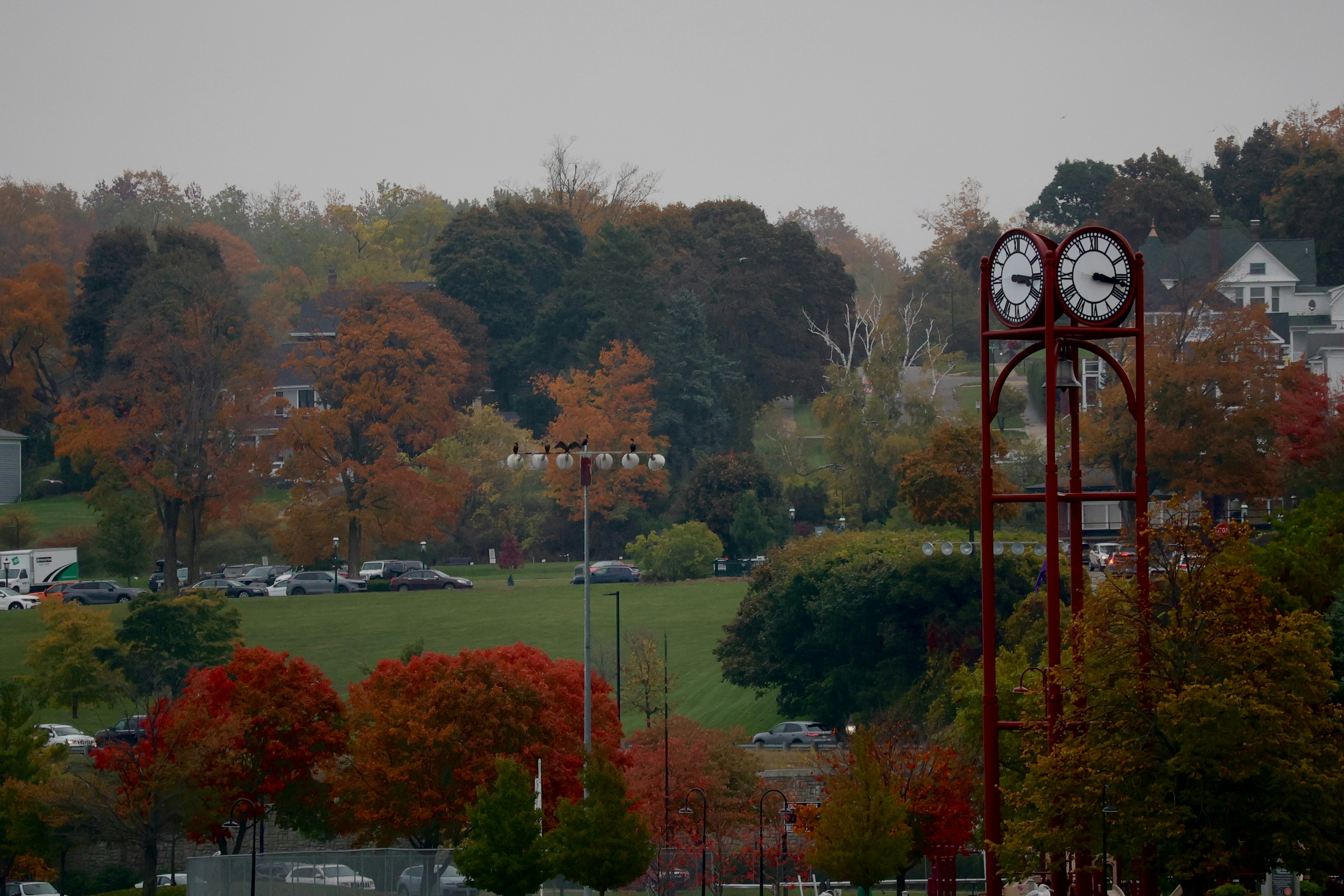 Autumnal park scene with a red clocktower standing amidst colorful trees; distant houses and a grassy park provide context and depth.