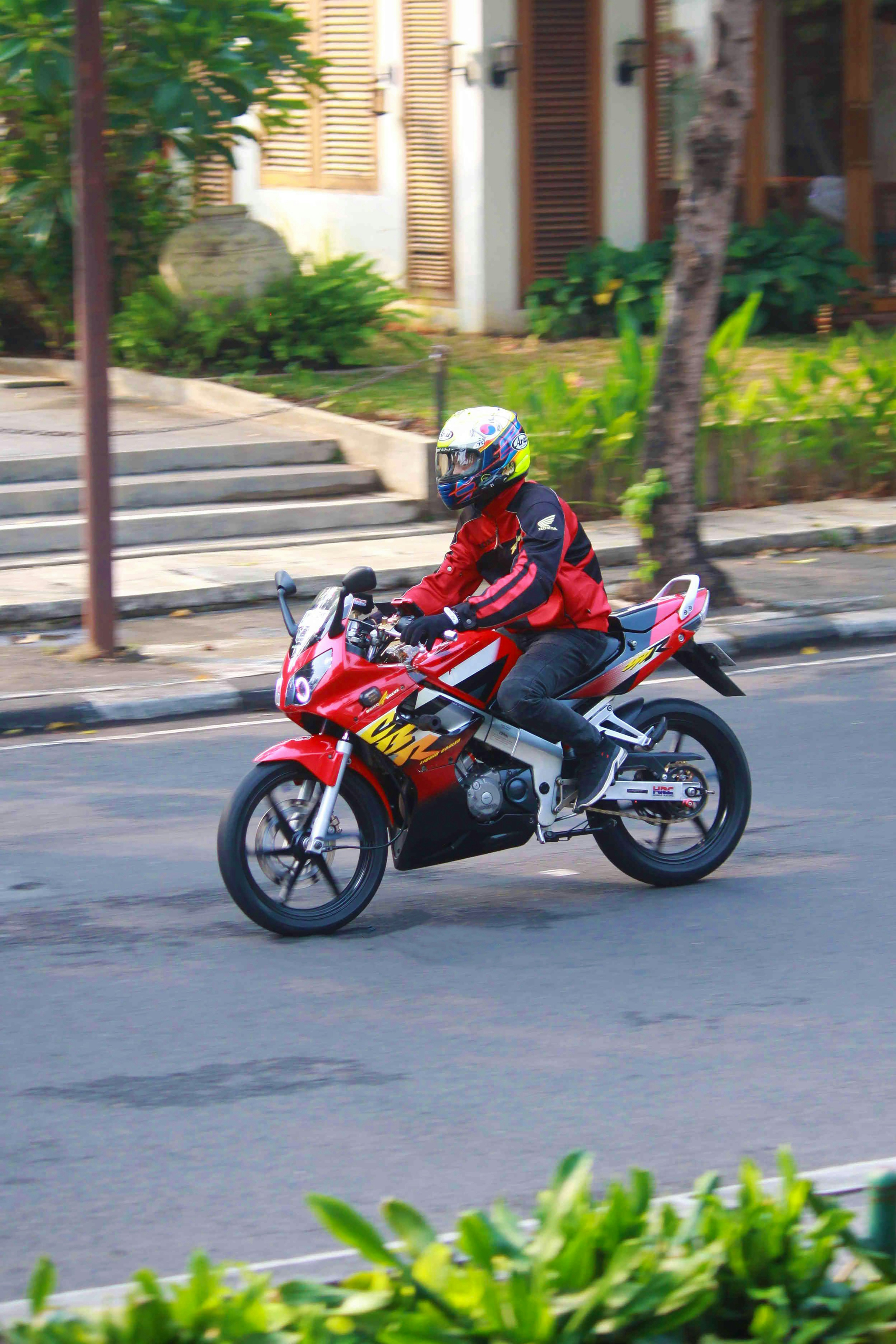 A motorcyclist in red gear rides a red sport bike along a calm street with greenery and houses in the background. The image captures a sense of speed and street-scene motion.