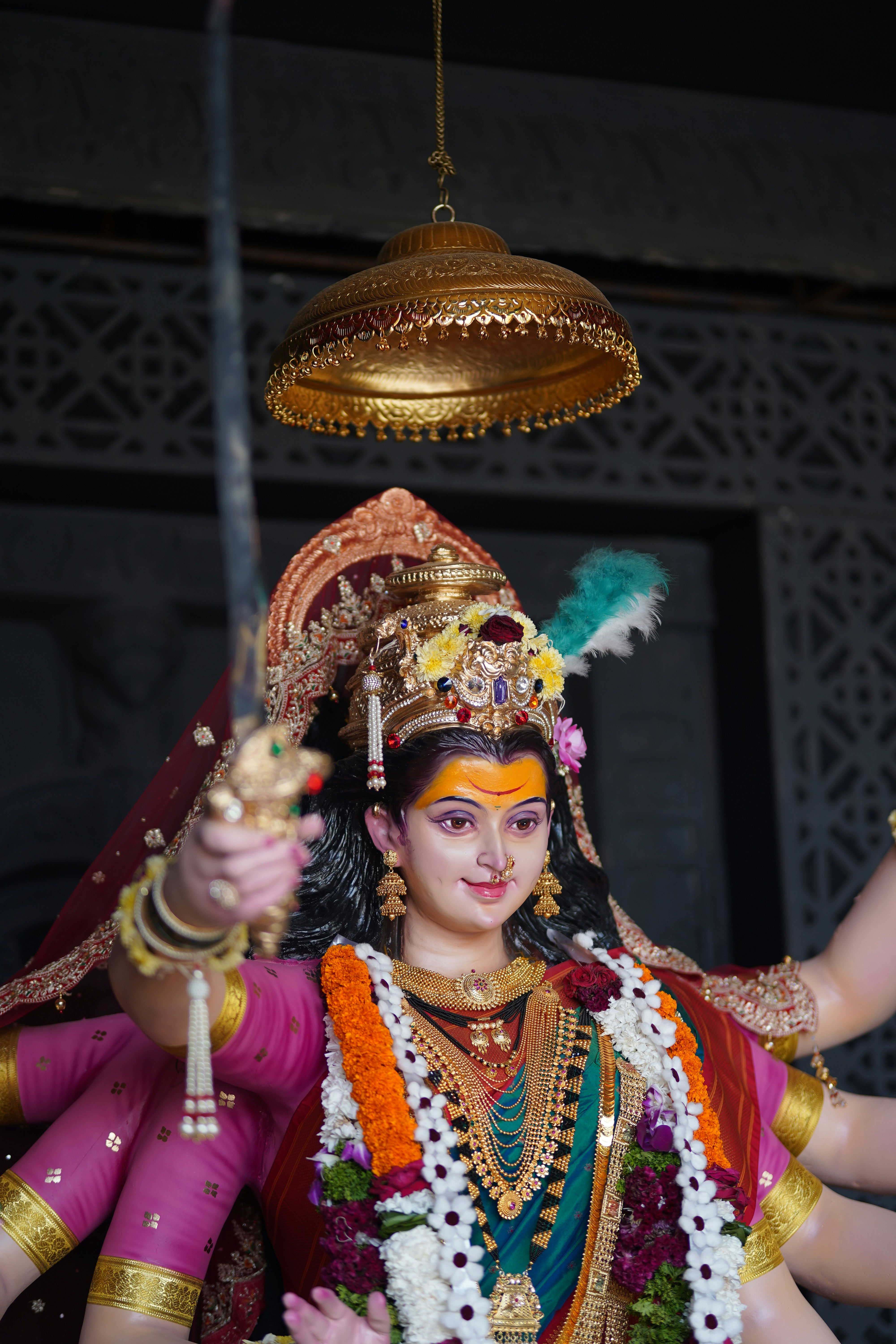 Colorful dancer in elaborate temple attire with intricate jewelry and floral garlands, mid-gesture under a decorative hanging lamp.