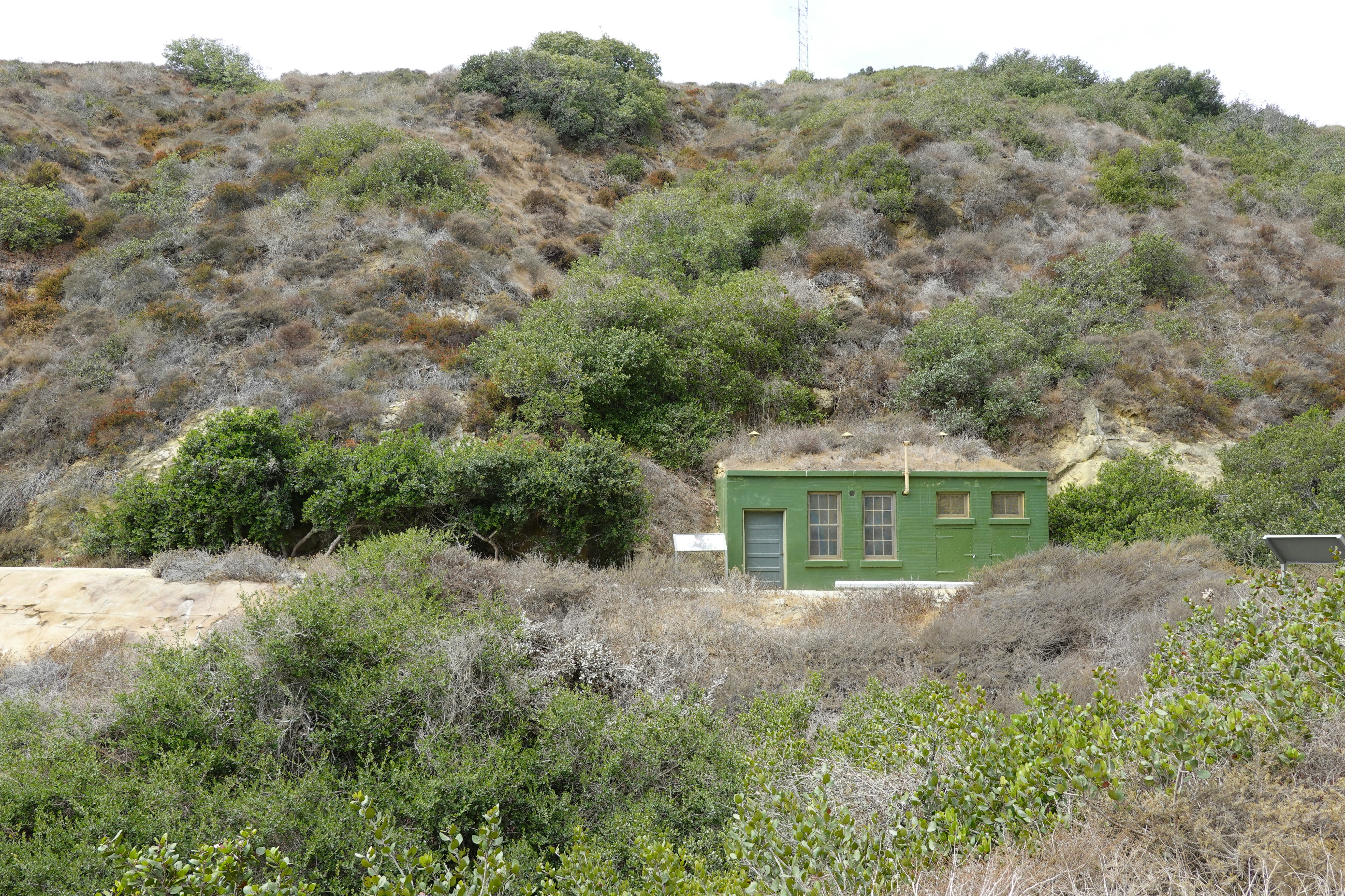 Small green cabin nestled among dry hillside shrubs and scattered trees; a quiet rural outpost with muted natural tones.