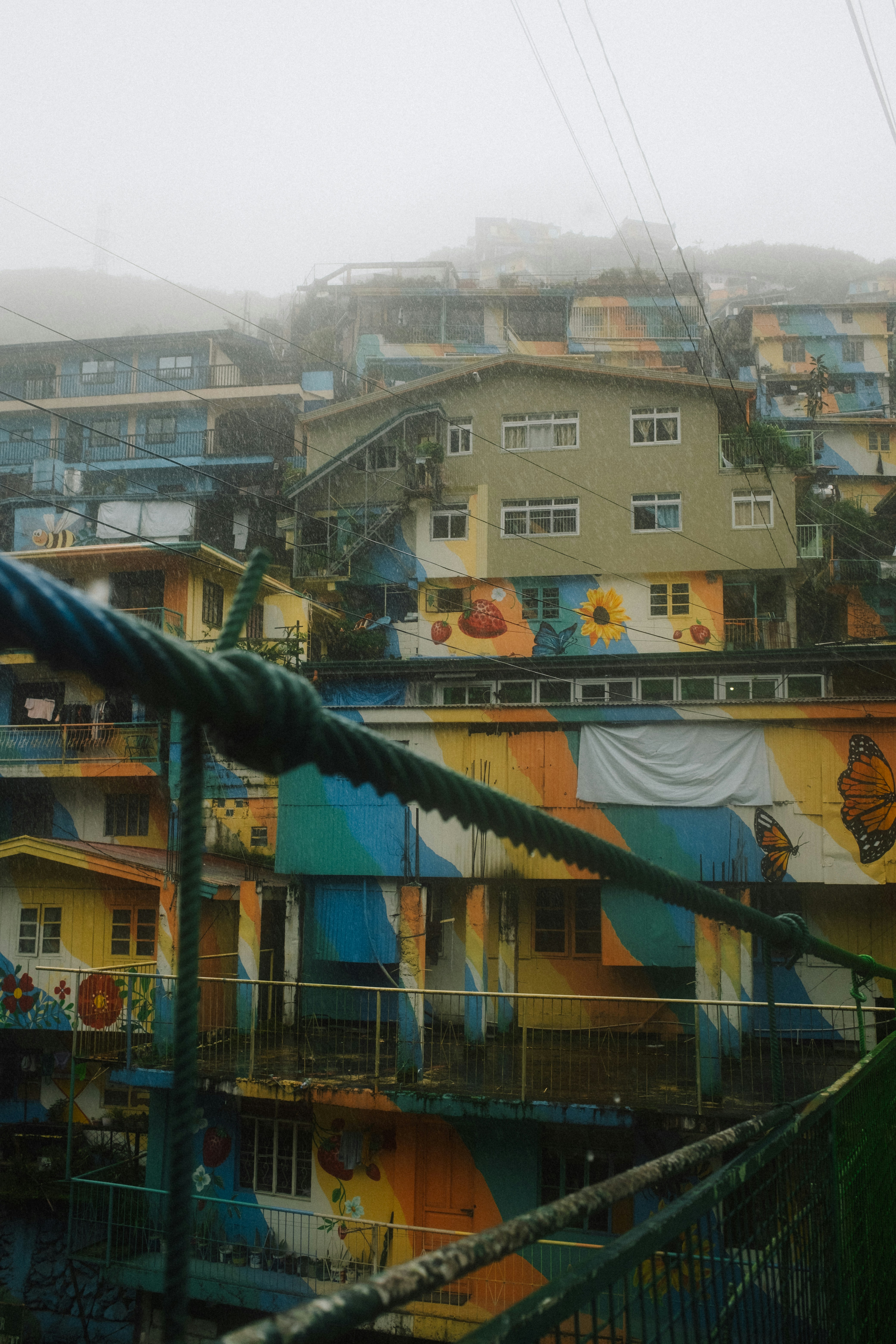 Colorful hillside apartments under mist with painted murals and a rain-soaked railing