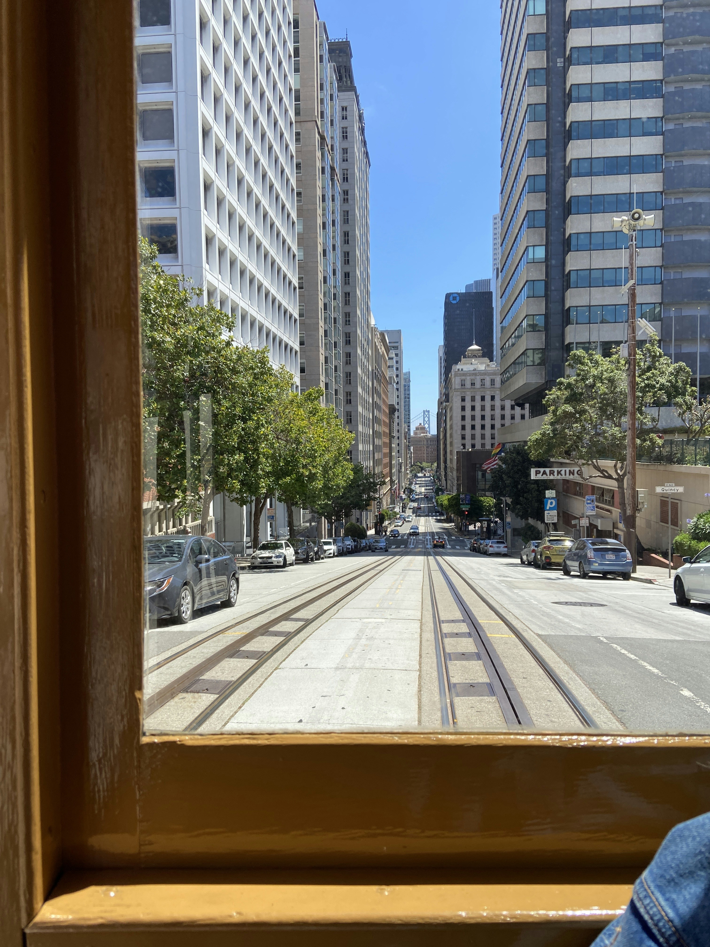City street viewed through a wooden frame, with tram tracks leading into the distance between tall buildings and parked cars along the curb.