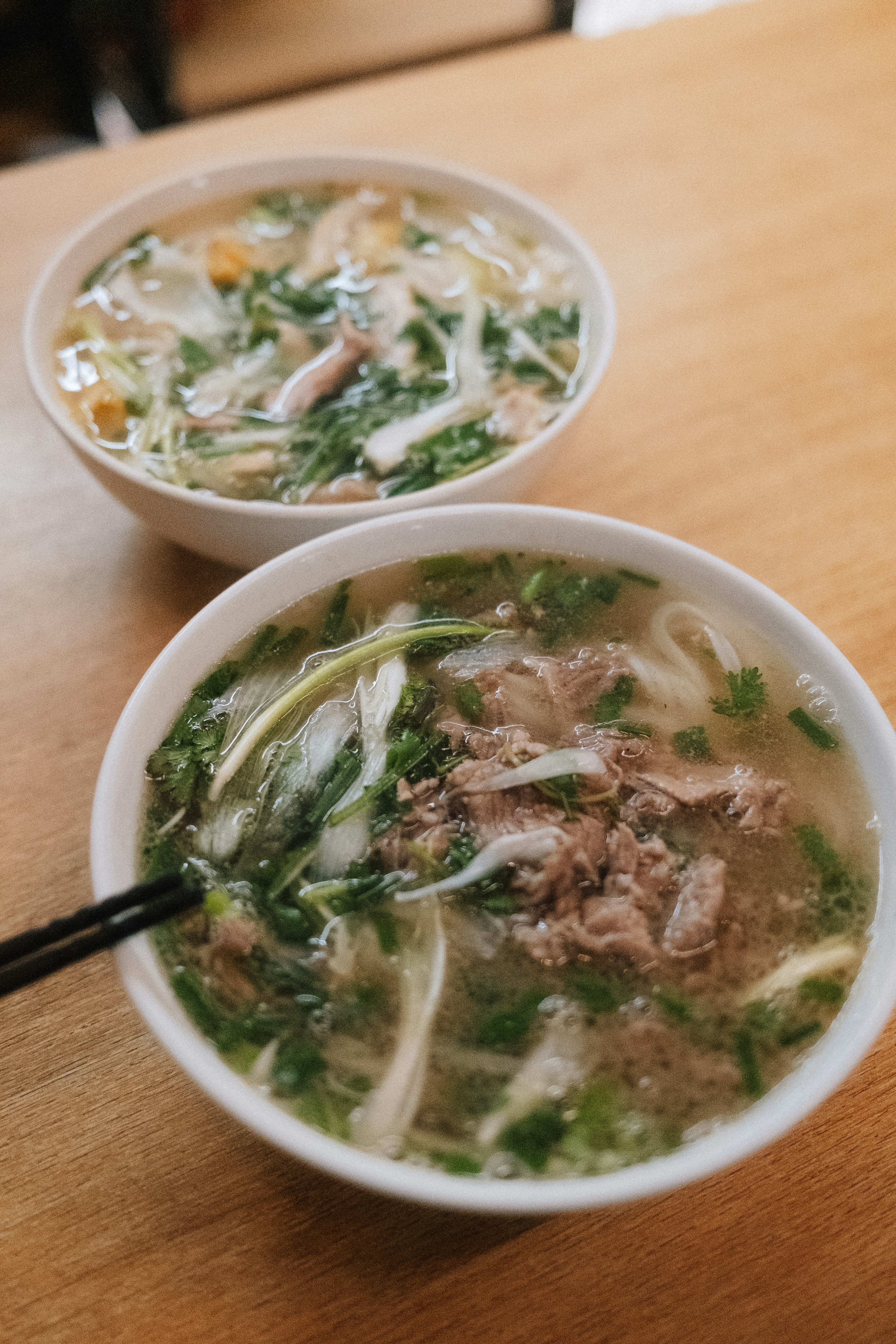Two bowls of steaming beef and herb noodle soup on a wooden table