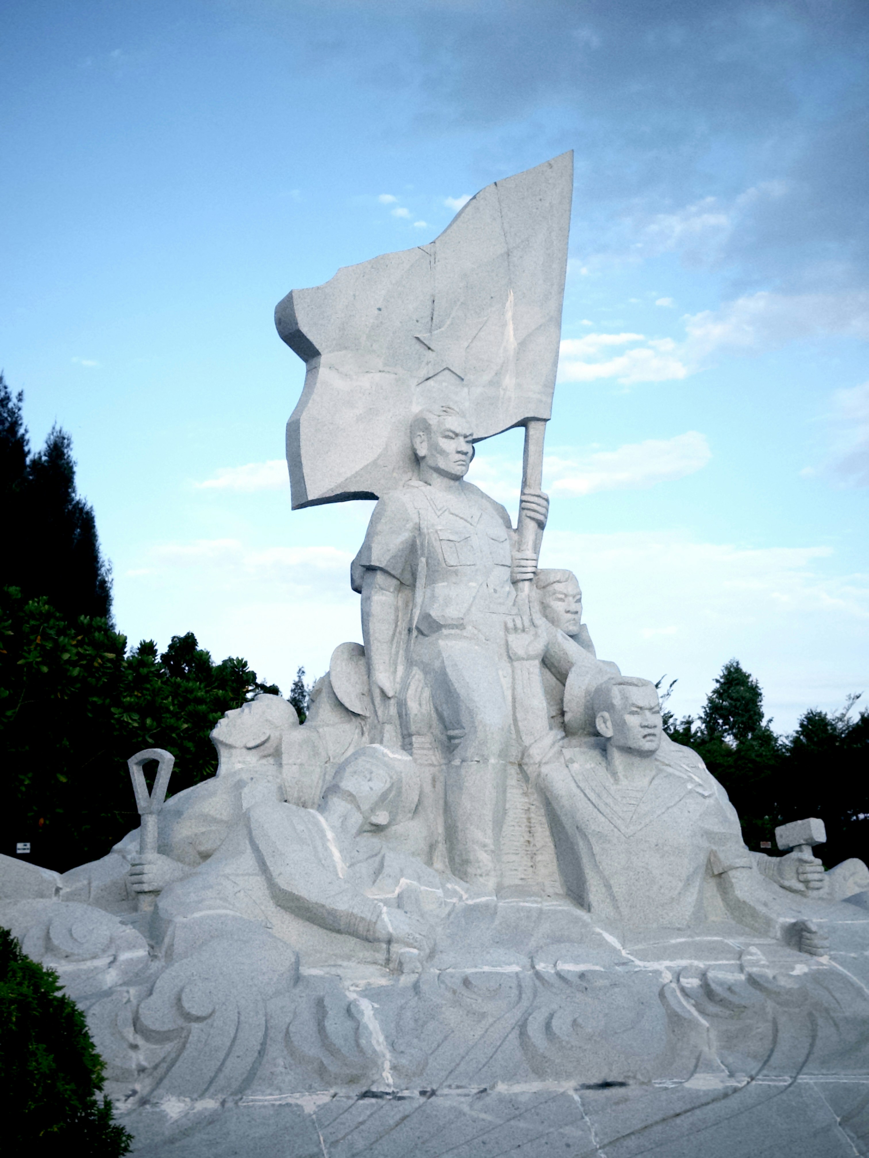 Monumental stone sculpture with multiple figures and a raised flag under a clear blue sky, carved in a dramatic, sweeping base. This is a static sculpture, not a painting or photograph.