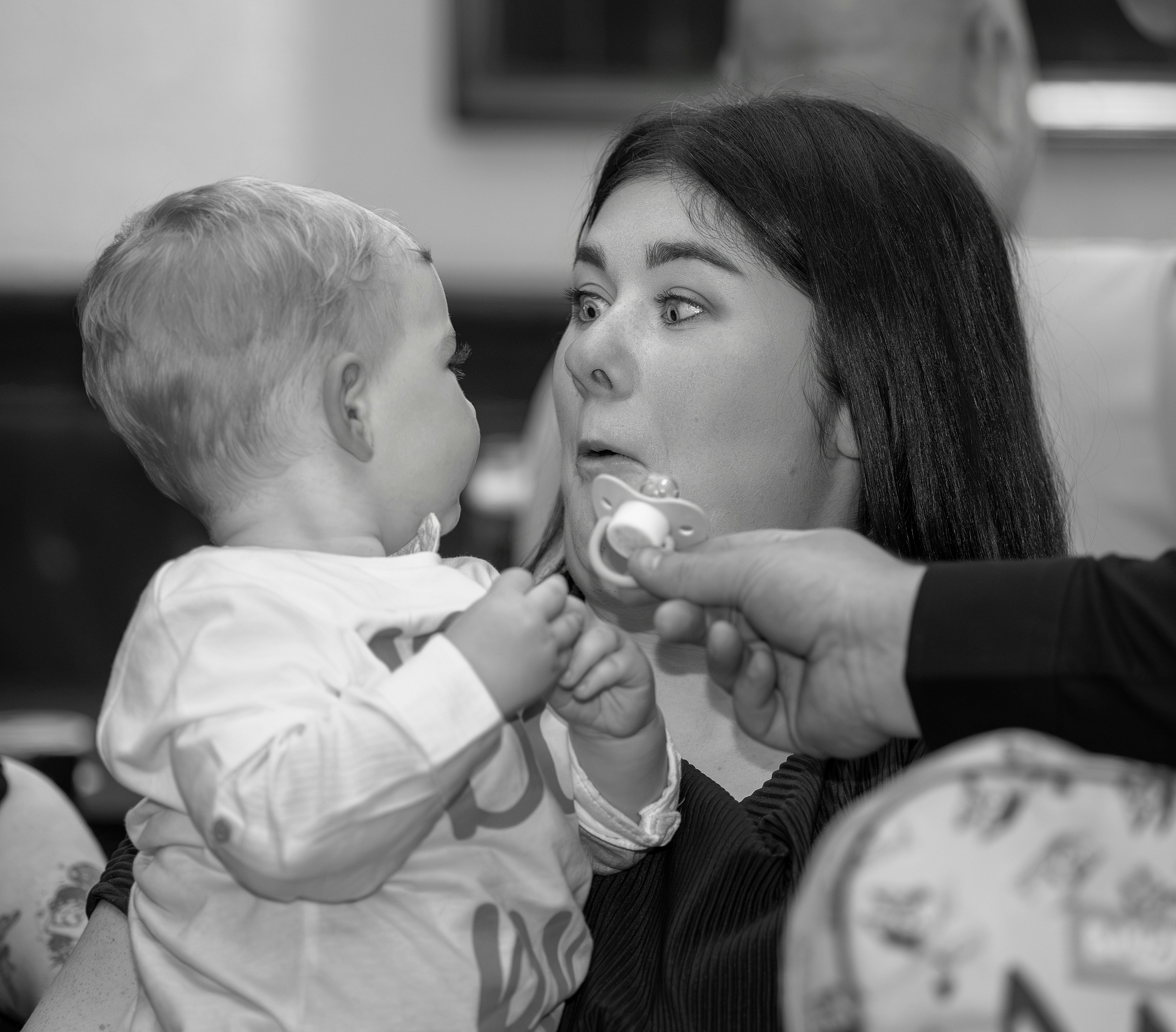 Mother with dark hair offers a pacifier to a baby held facing her in a candid moment inside a home.