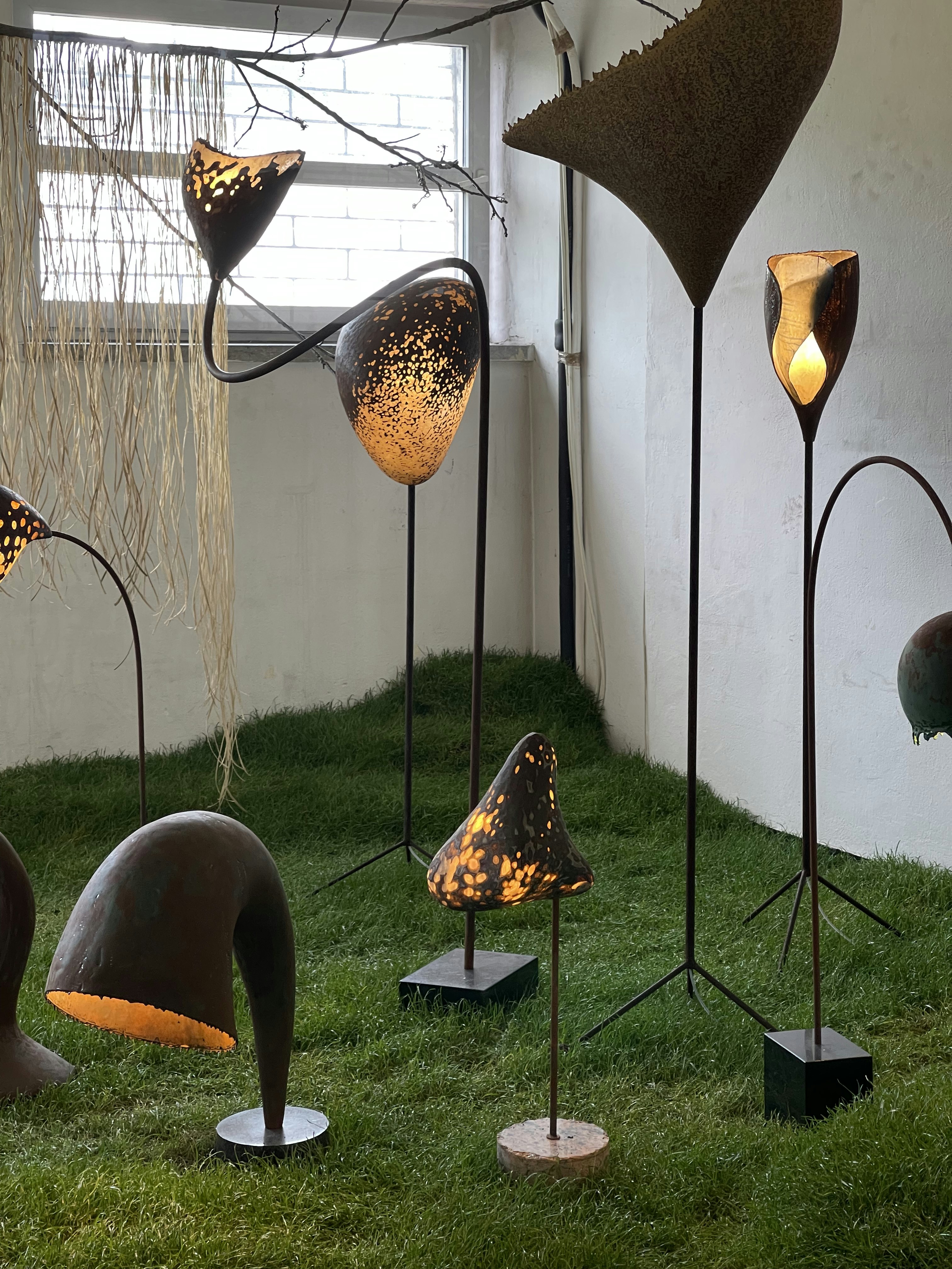 An indoor installation of rusted metal sculptures on a green carpet, with a bright window in the background; the scene emphasizes industrial forms and warm light from perforated surfaces.