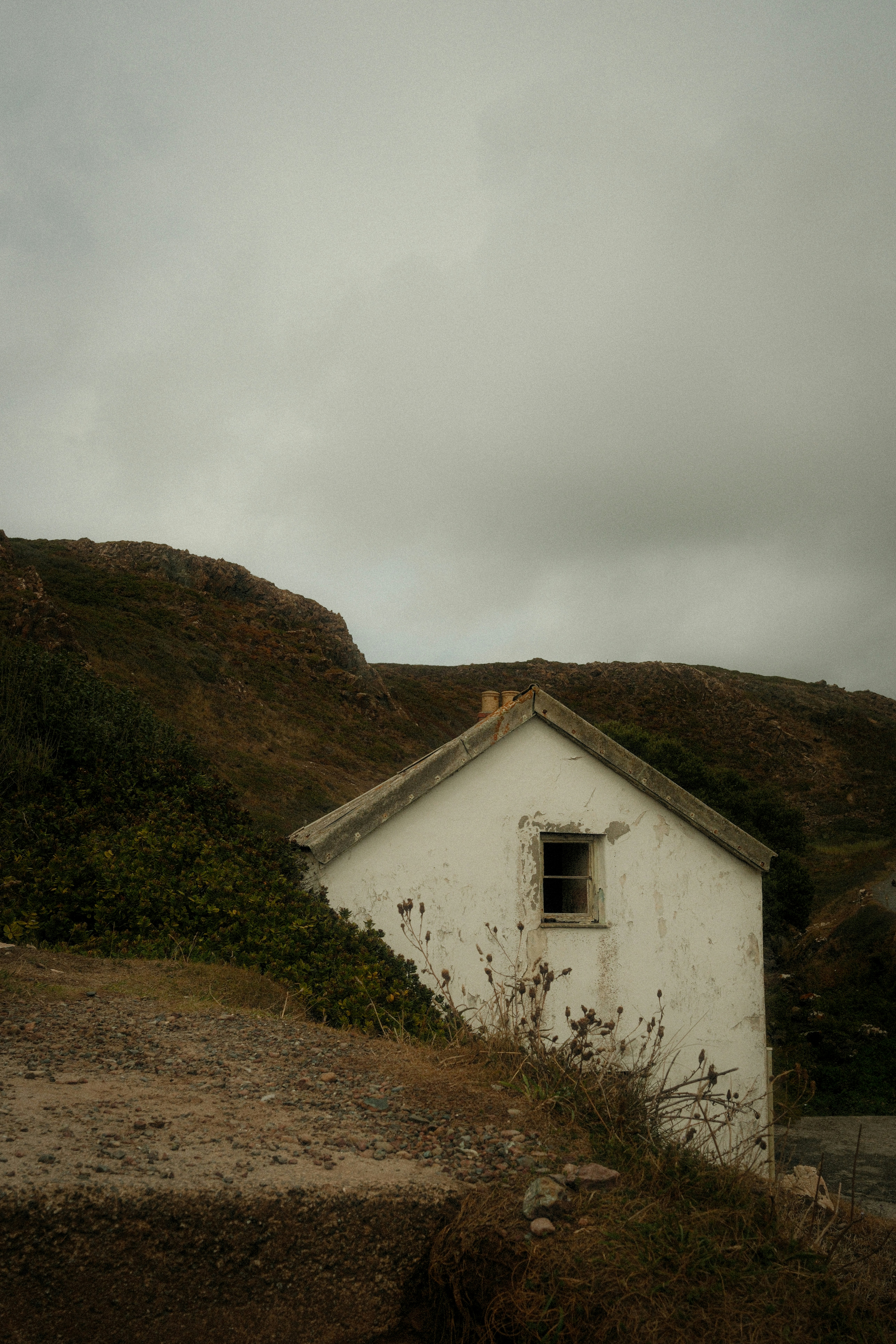 Sparse hillside scene with a solitary white house perched near a dirt foreground and scrubby vegetation under a cloudy sky. A simple architectural subject against rugged landscape.