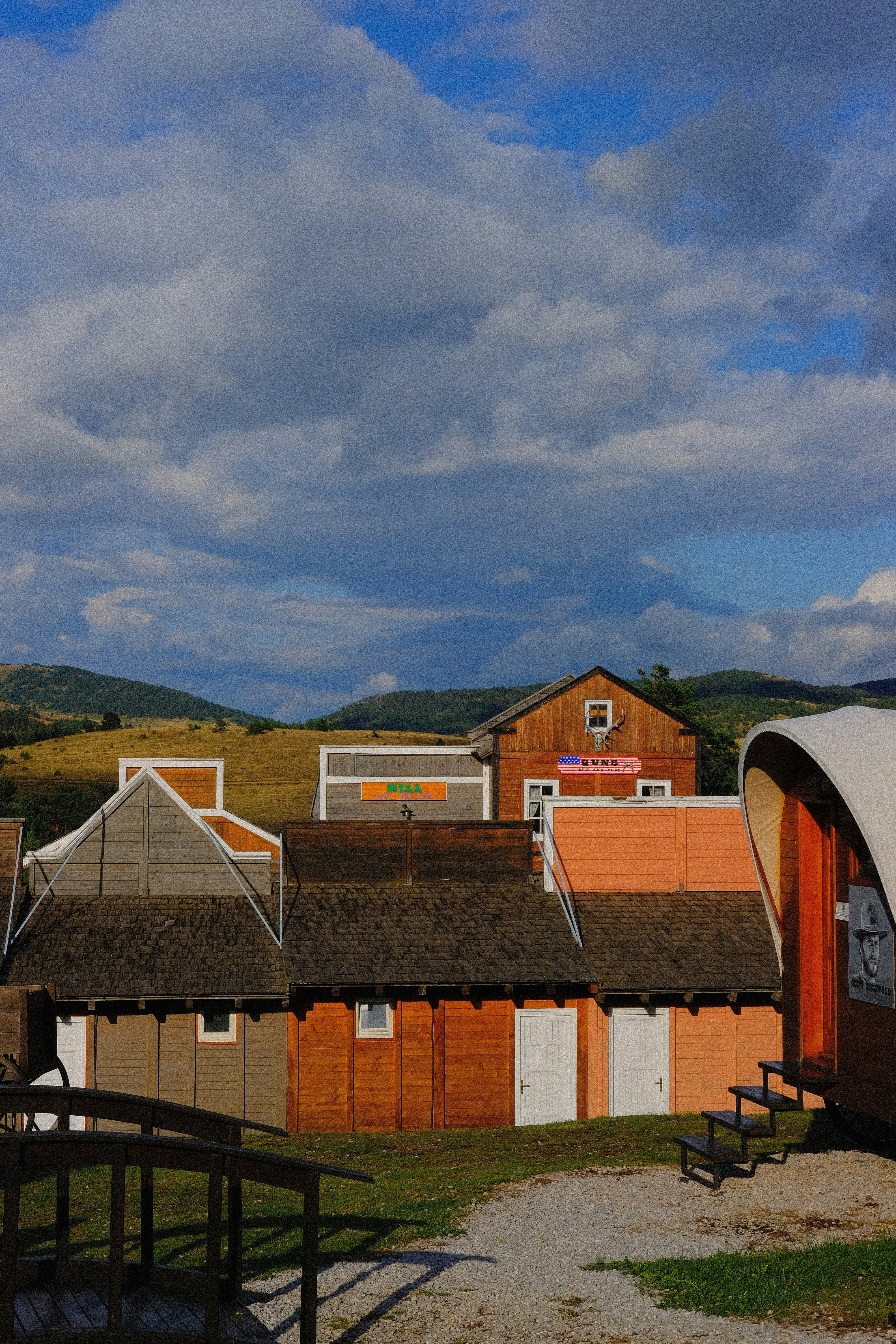Row of rustic cabins and storefronts along a gravel path under a dramatic cloudy sky; foreground includes steps and a curved shelter, suggesting a rural or historical market setting.