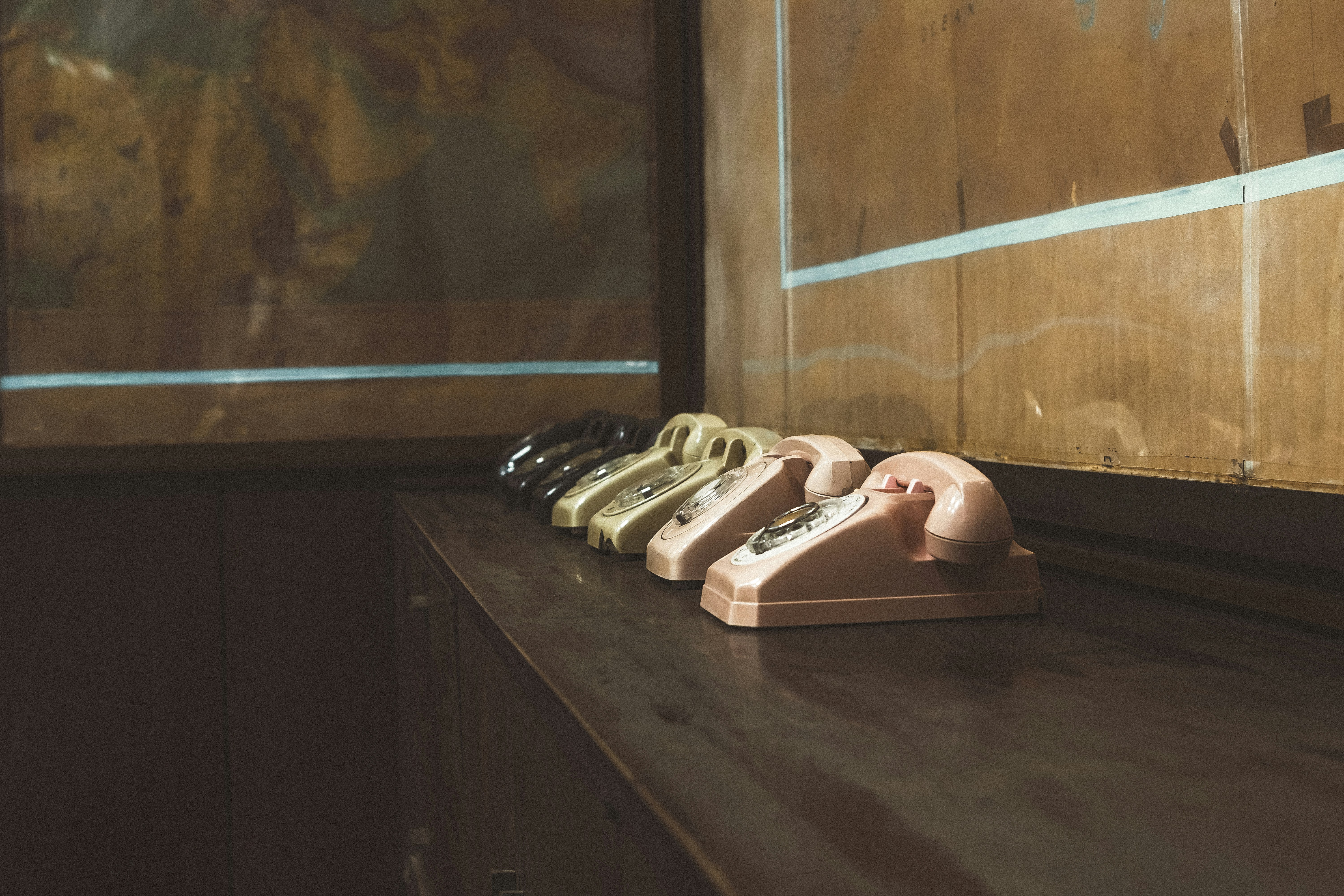 Muted retro telephones sit in a line along a dark wooden counter, with a textured wall backdrop and soft, moody lighting.