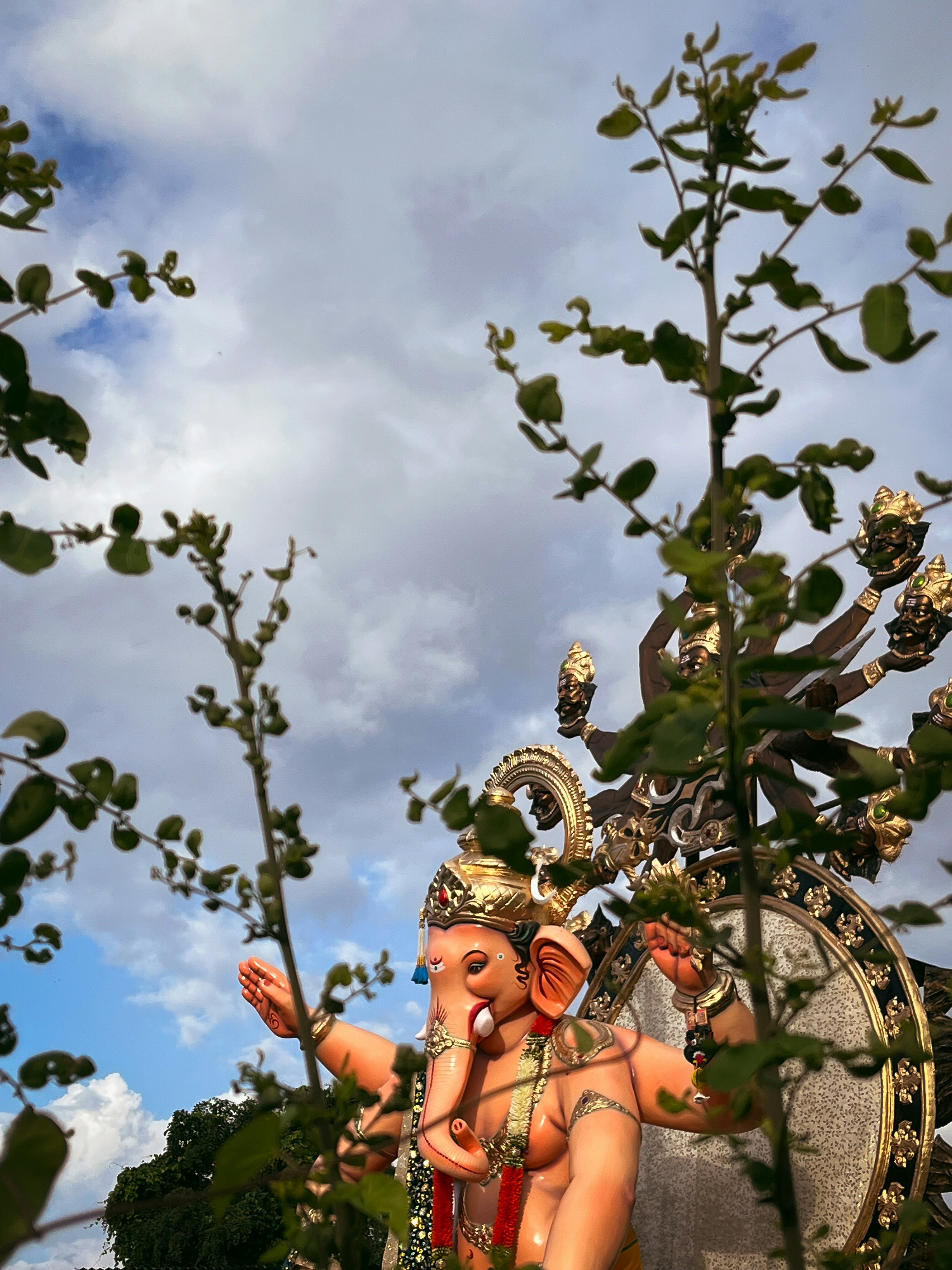Close-up view of a decorative Ganesha statue partially framed by foreground branches against a blue sky, highlighting intricate details and festive adornments.