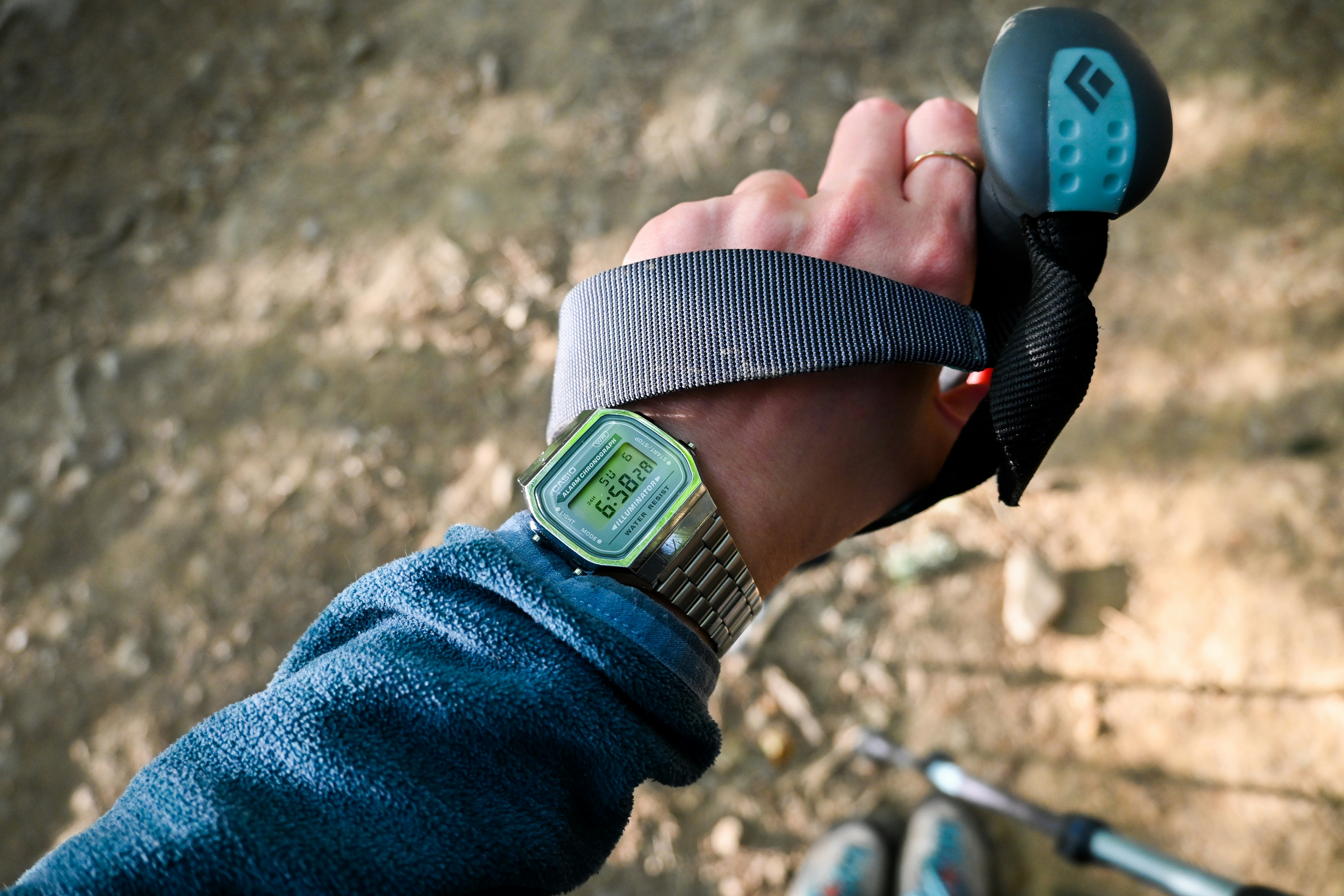 Close-up of a wrist wearing a digital watch during a climb, with a harnessed grip and a blurred outdoor trail in the background. This is a candid action shot emphasizing gear and readiness.