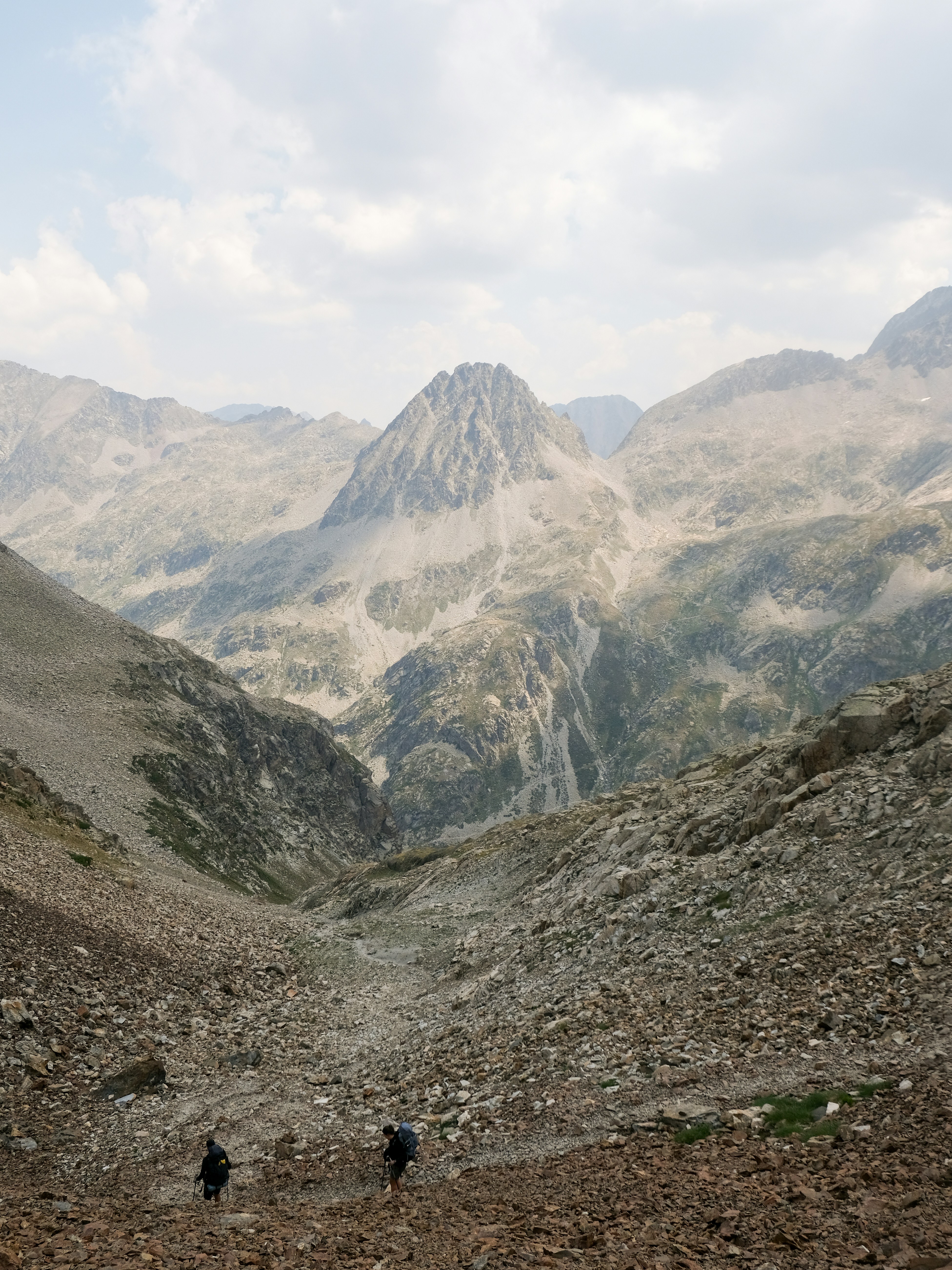 Two climbers descend a rocky alpine valley toward jagged peaks under a pale sky, emphasizing rugged terrain and scale.