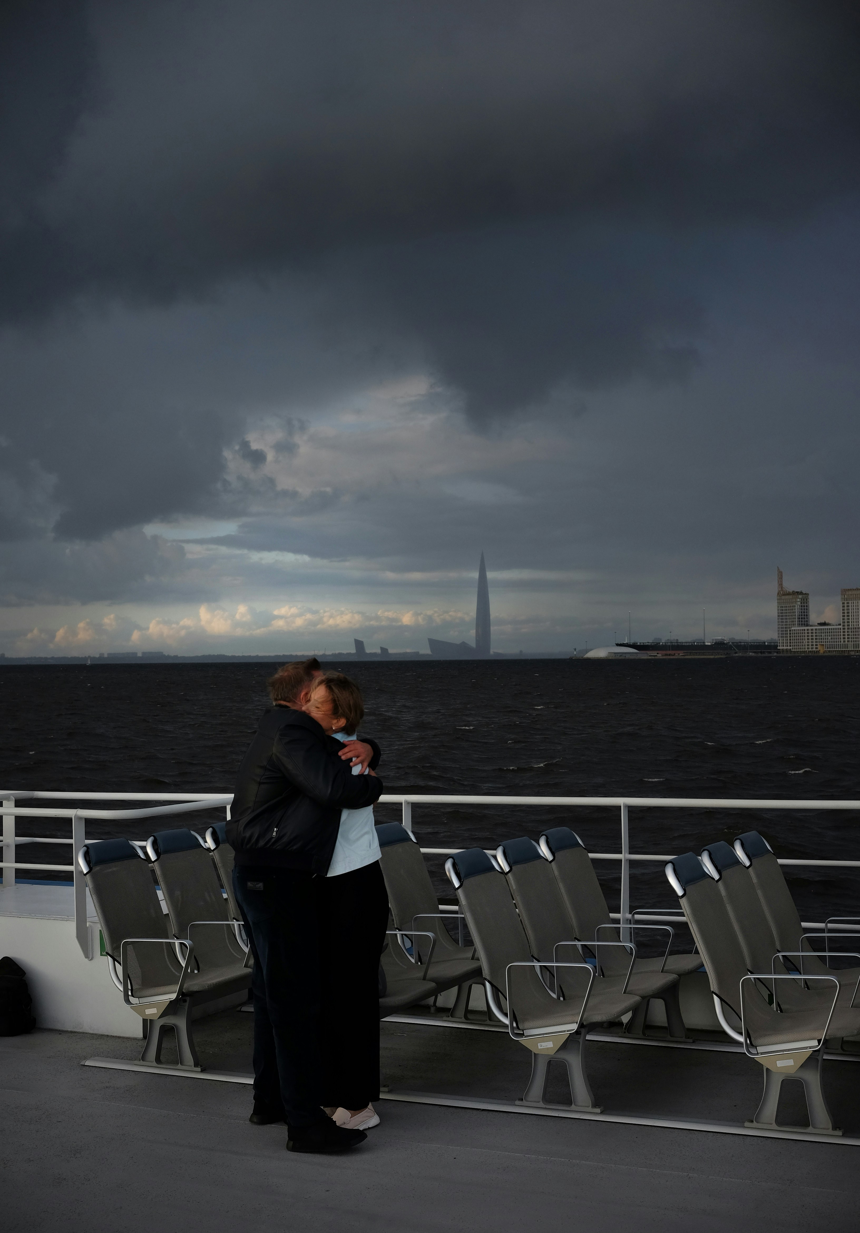Two people share a warm kiss on a windswept pier with a distant city skyline and dark storm clouds overhead.