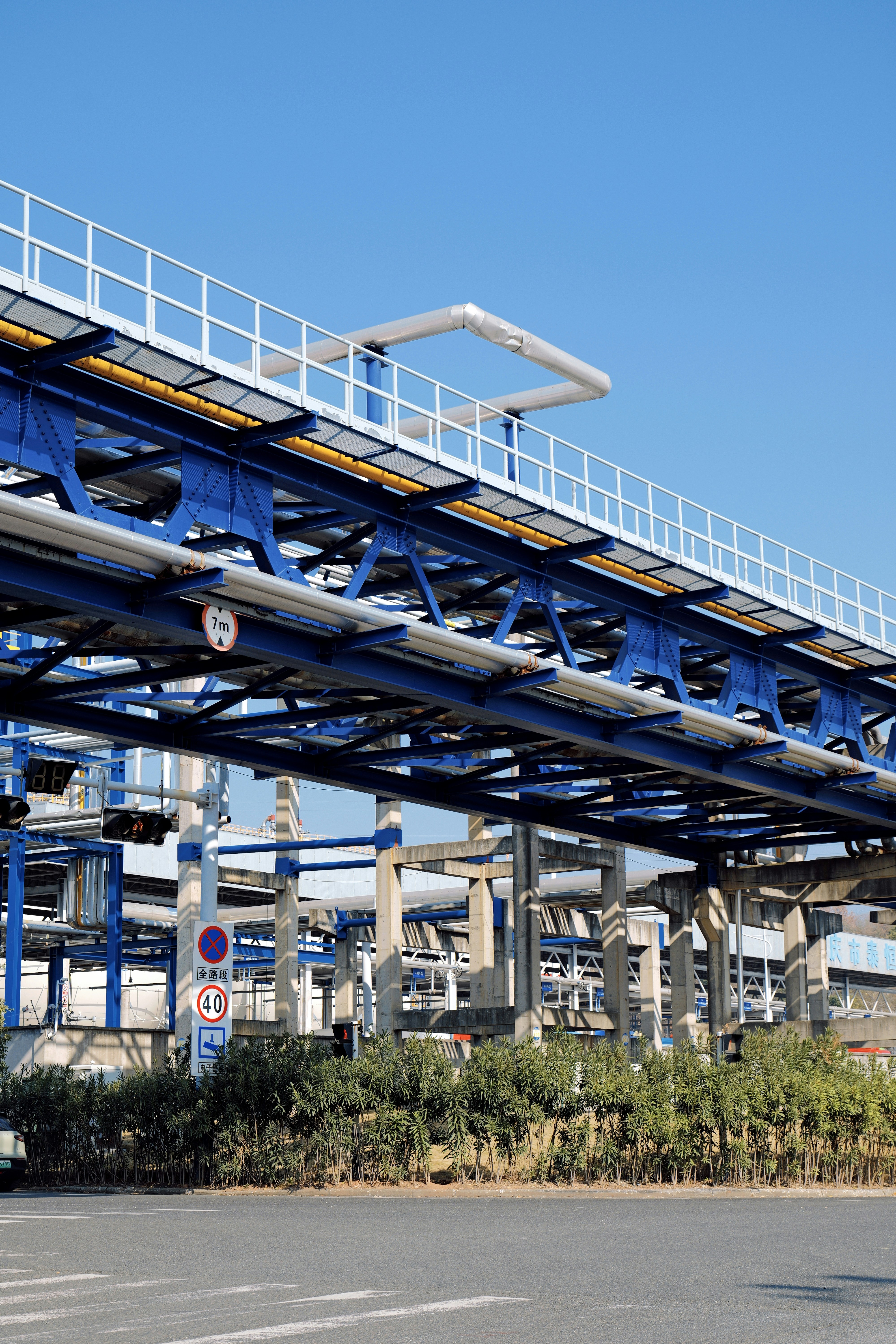 A blue industrial pipe bridge over a road with traffic signs and bushes