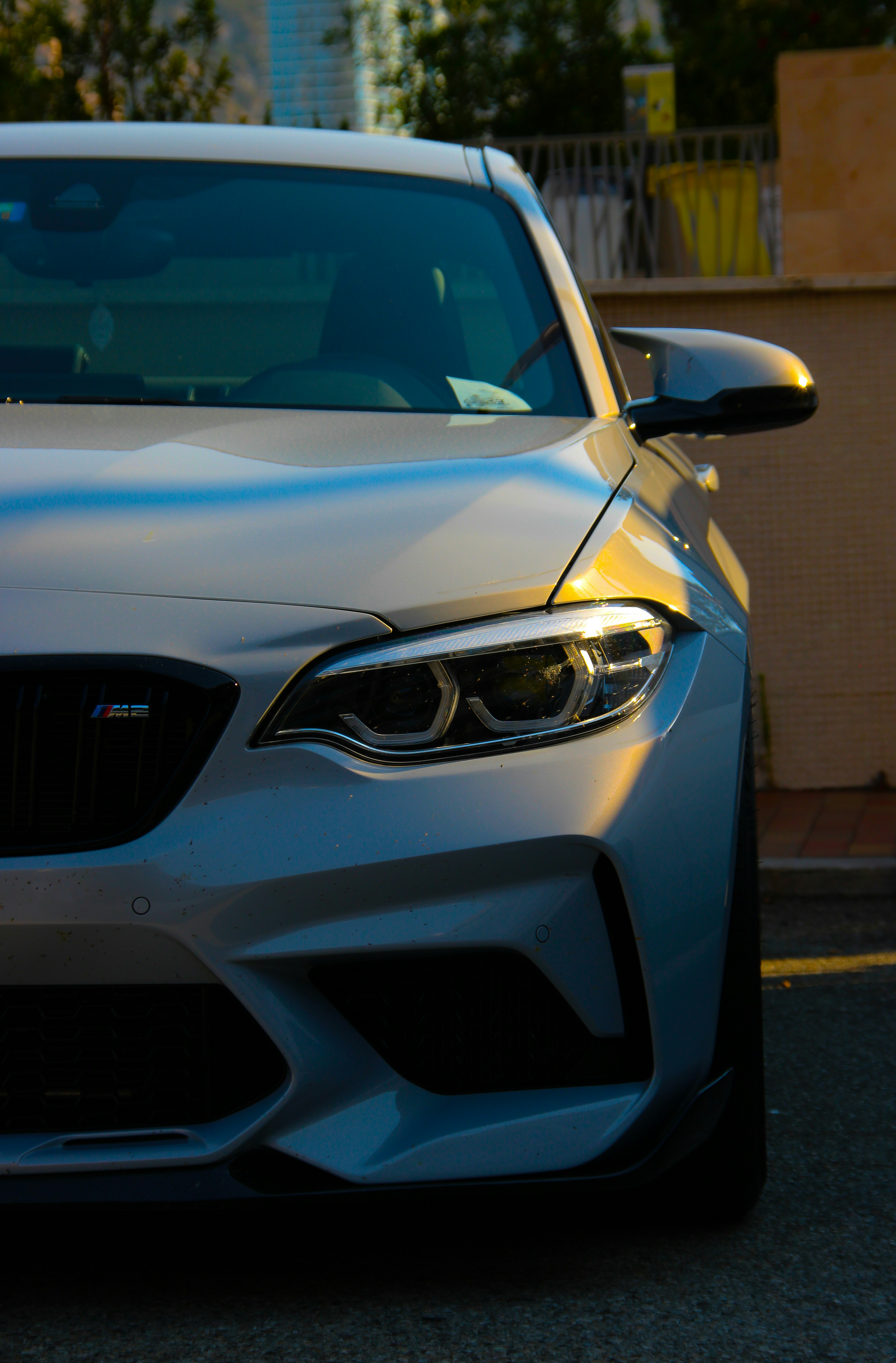 Close-up of the front-left section of a white BMW with distinctive headlight and aggressive lower fascia, parked in an urban lot. The image emphasizes the car's sculpted lines and glossy finish.