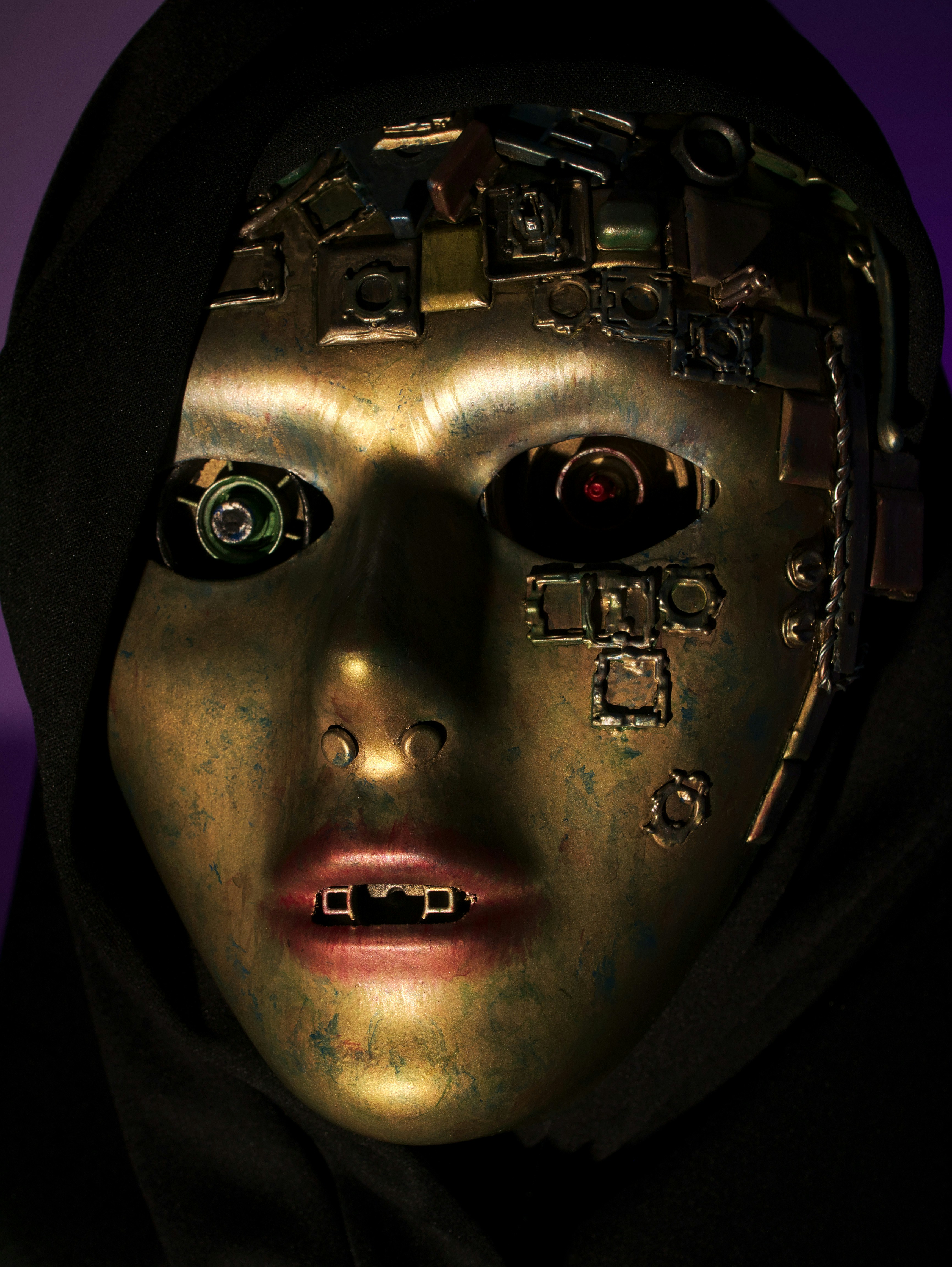 A close-up portrait of a metallic, partially deconstructed mask under dark lighting, with a hood framing the face and subtle purple backlight. This is a sculpture-like photograph emphasizing texture and concealed features.