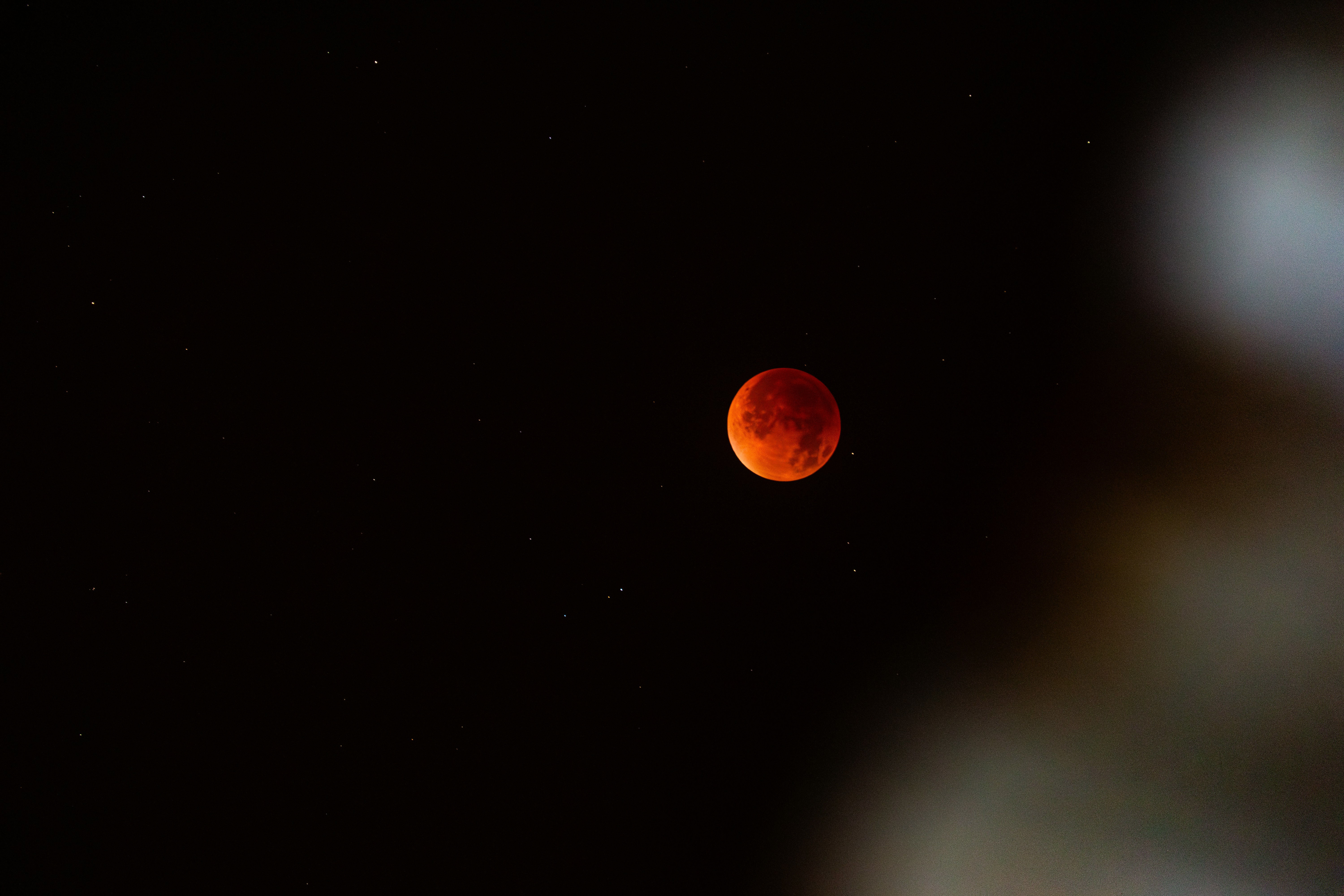 Distant blood-red moon amid dark skies with scattered stars and a softly blurred foreground edge. The scene emphasizes isolation and celestial calm.