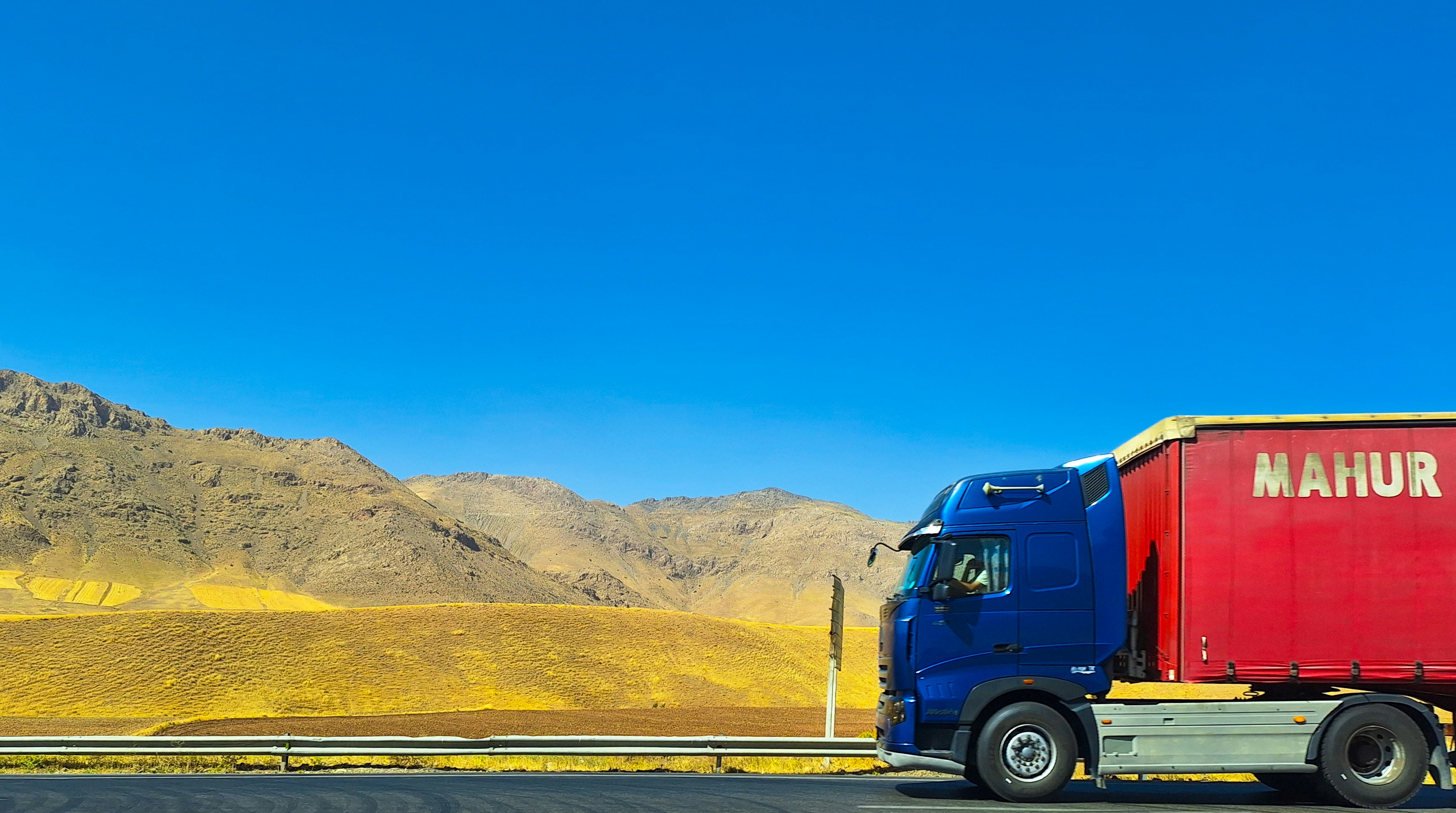 Blue semi-truck on a barren desert highway beneath a clear blue sky; long red trailer contrasts with arid landscape and distant mountains.