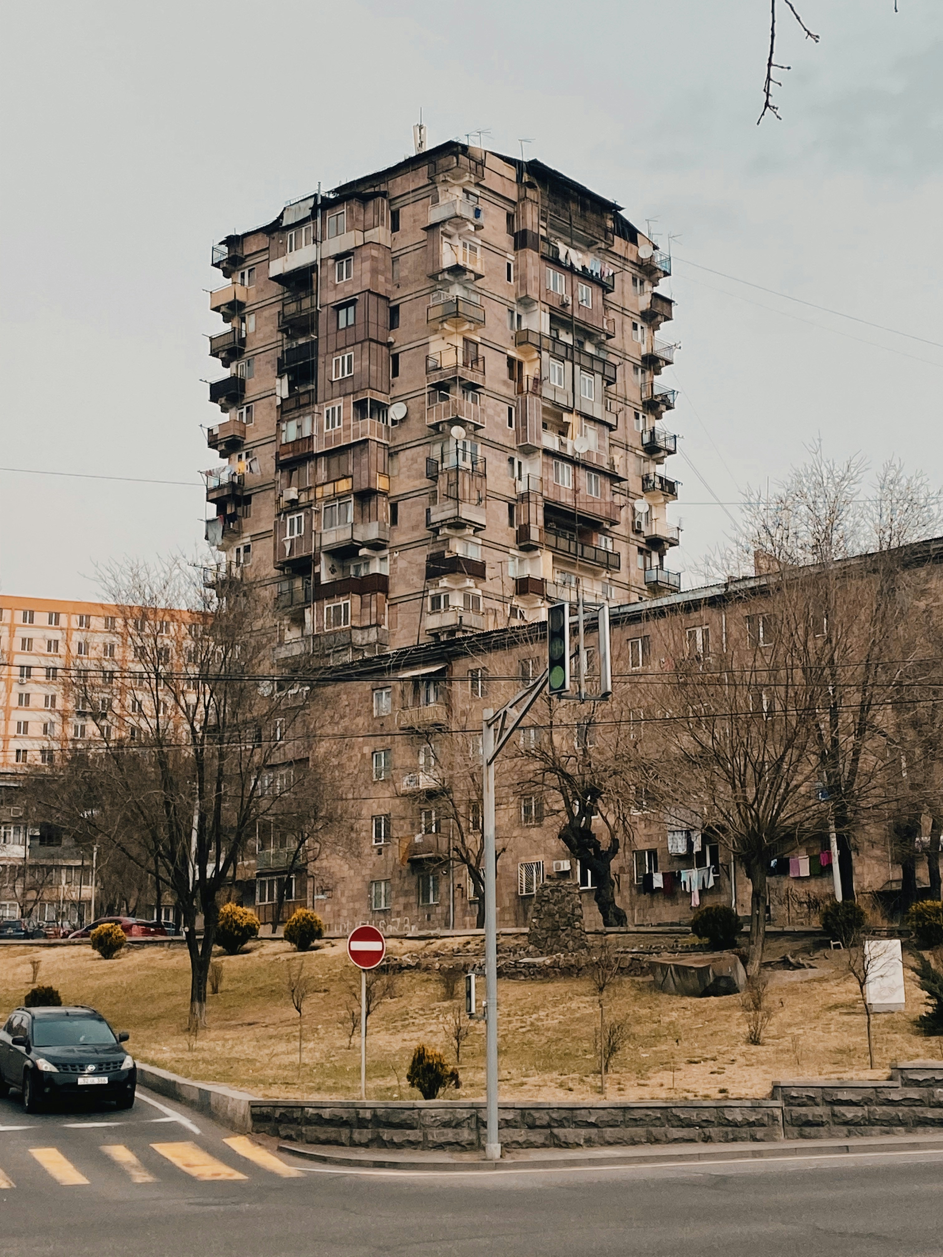 A tall, weathered brick apartment building dominates the scene with bare trees and a small park at its base, while a street with a pedestrian crossing and a car sits in the foreground.