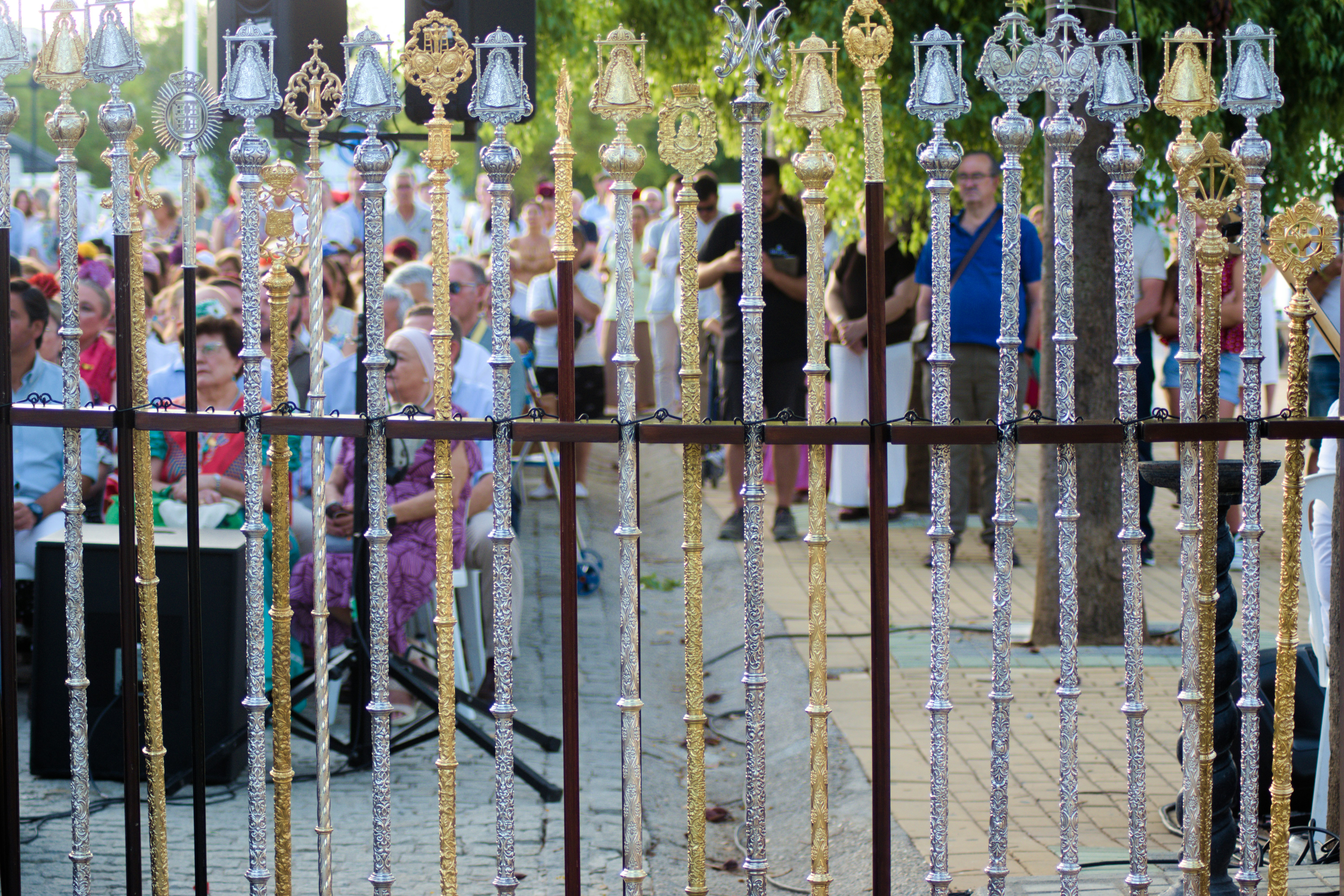 Crowd gathered behind a decorative iron fence, with ornate gold and silver posts creating a patterned barrier; a ceremonial or public event unfolds beyond the bars with seated attendees and onlookers.