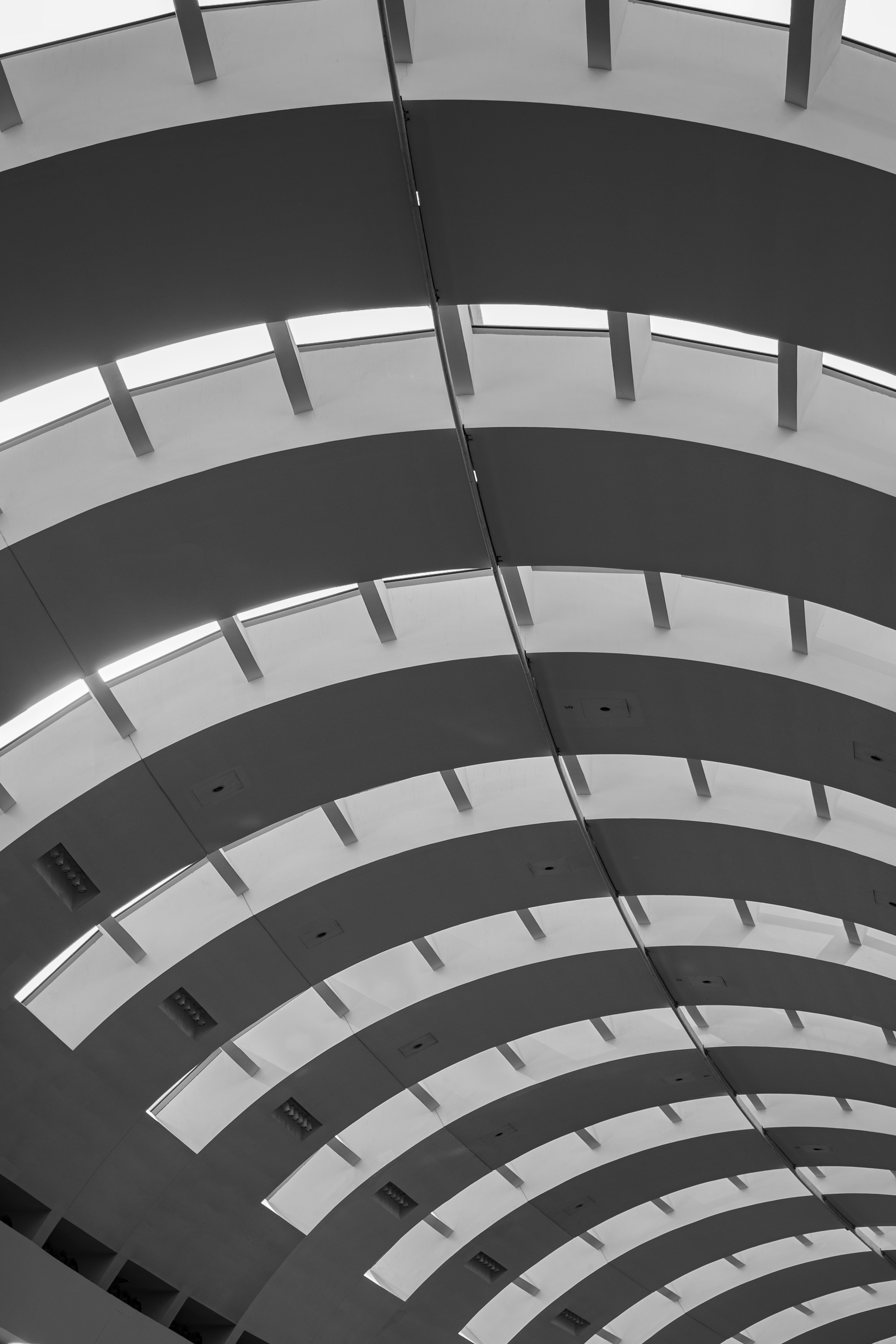 Monochrome view of a spiraling, concentric parking structure interior with alternating bands of light and shade; strong geometric repetition and perspective draw the eye toward the vanishing center.