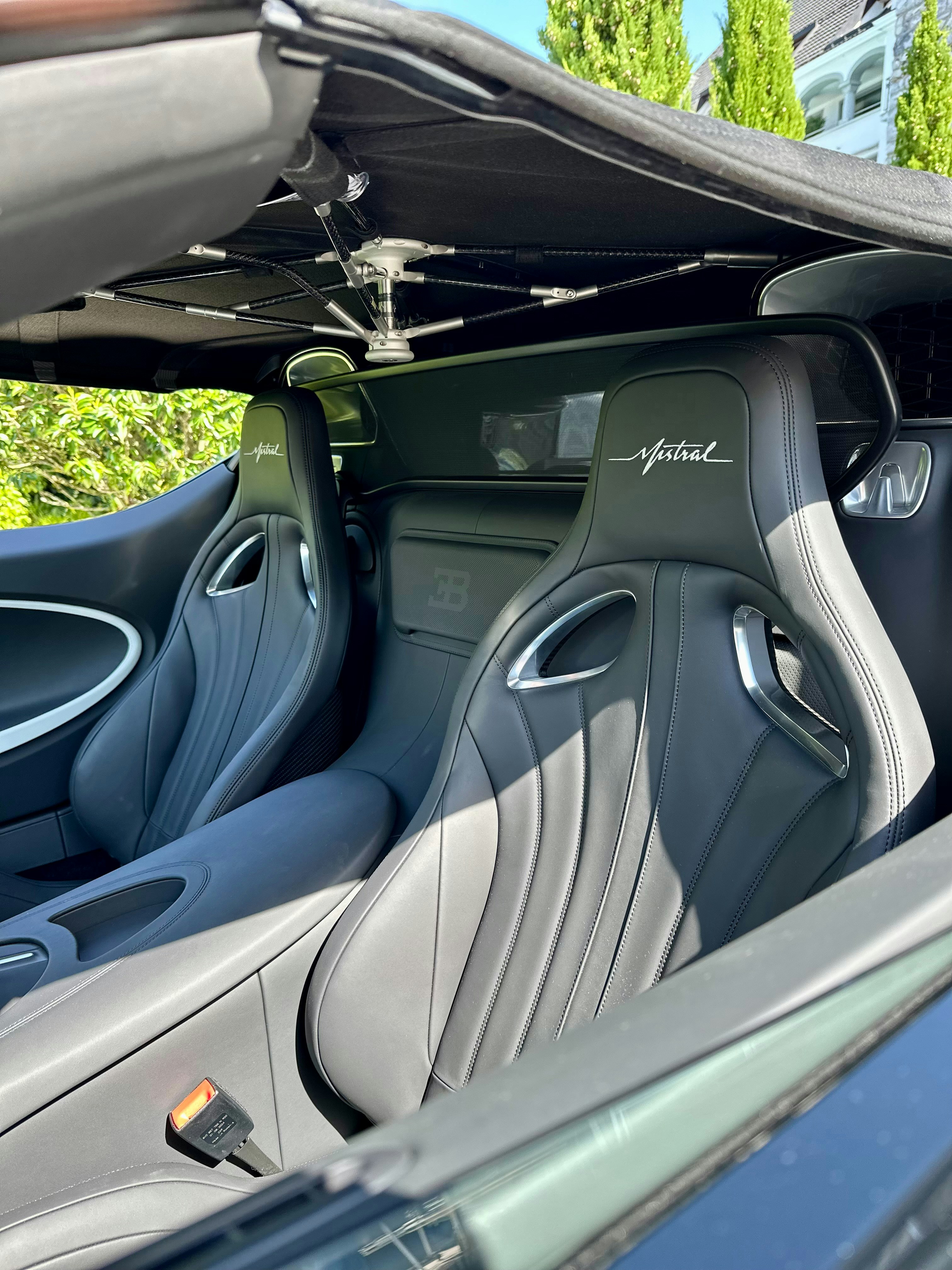 Luxury sports car interior with racing bucket seats and visible soft top mechanism; two sculpted seats branded with Mistral/branding, chrome accents, and exposed frame above.
