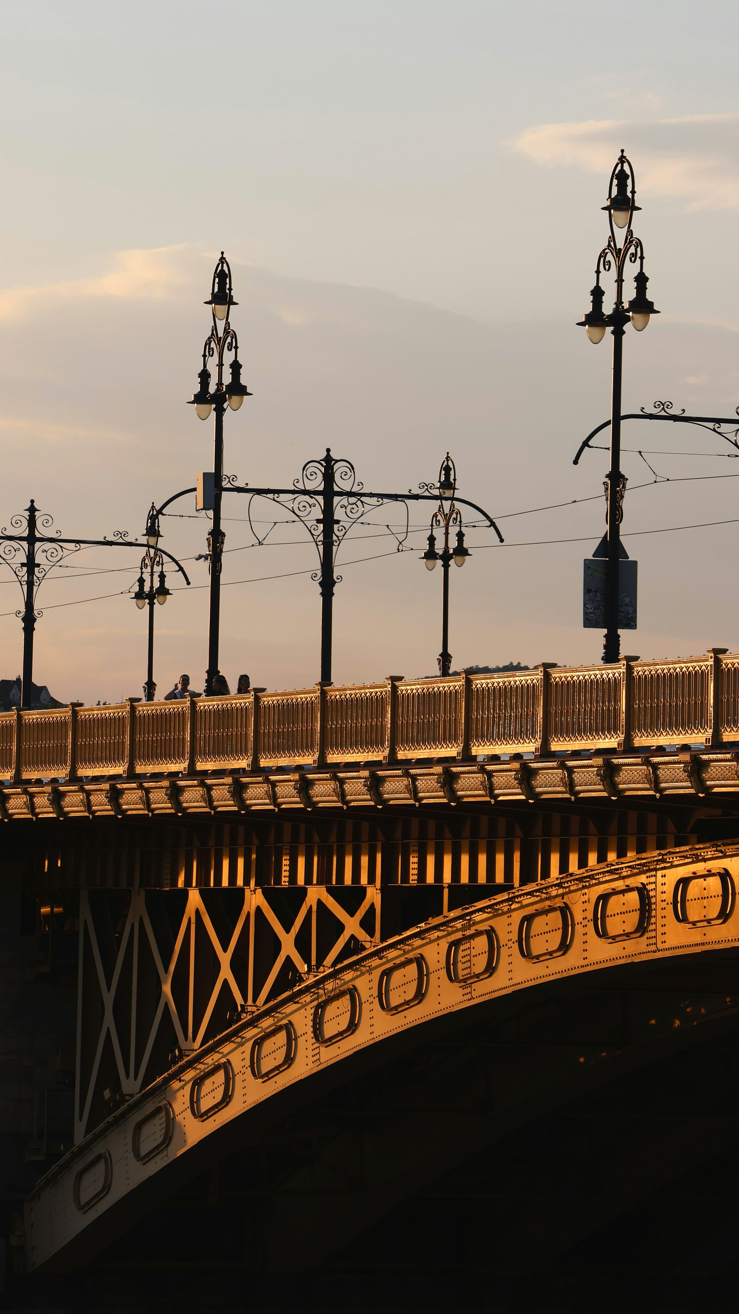 Sunset casts warm light over an ornate iron bridge, highlighting decorative lampposts and lattice railings along the railing. The scene emphasizes architectural detail and soft sky tones.
