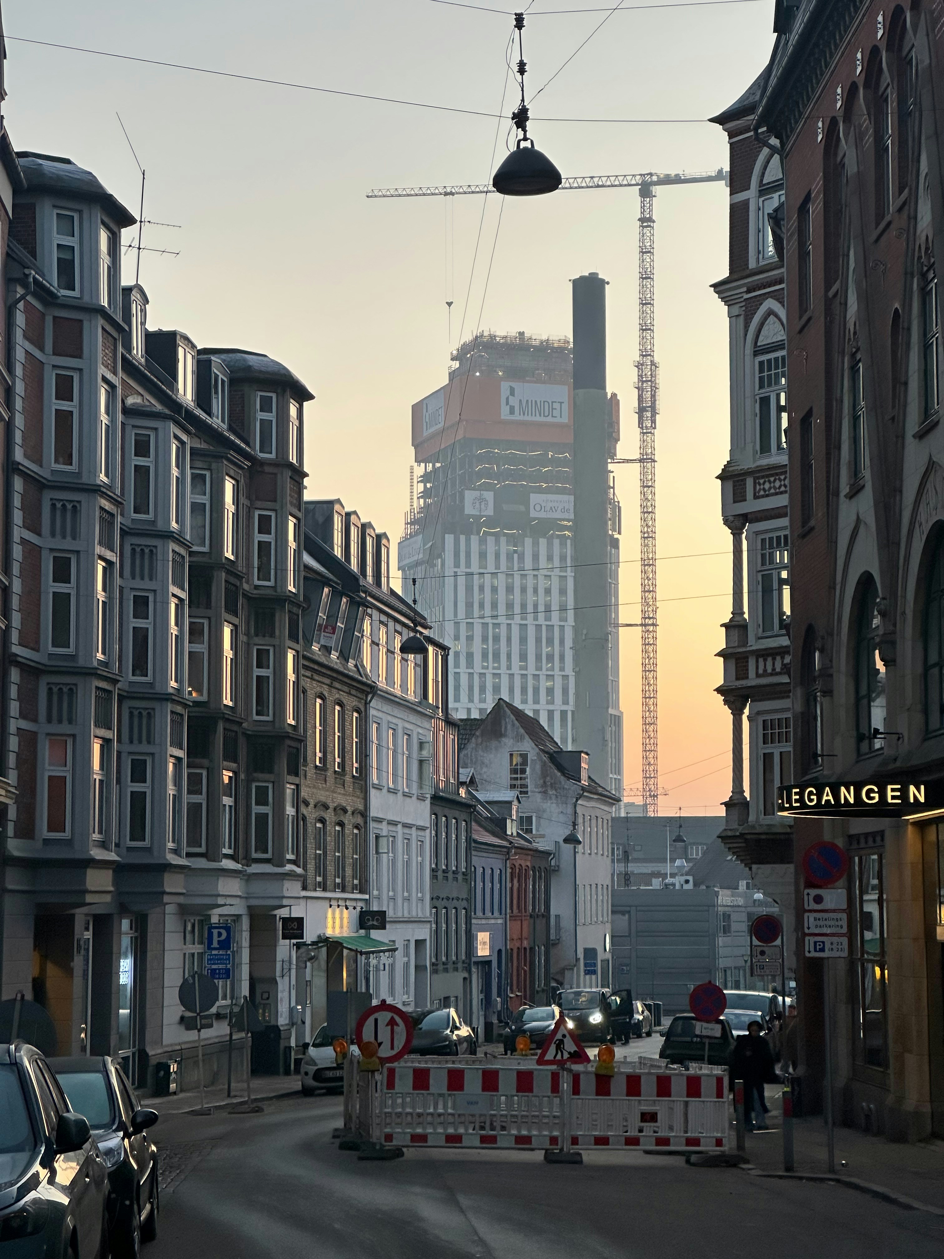 Dusk-lit city street lined with classic buildings leads to a tall construction site behind, with barrier blocks in the foreground and a partially visible shop sign on the right.