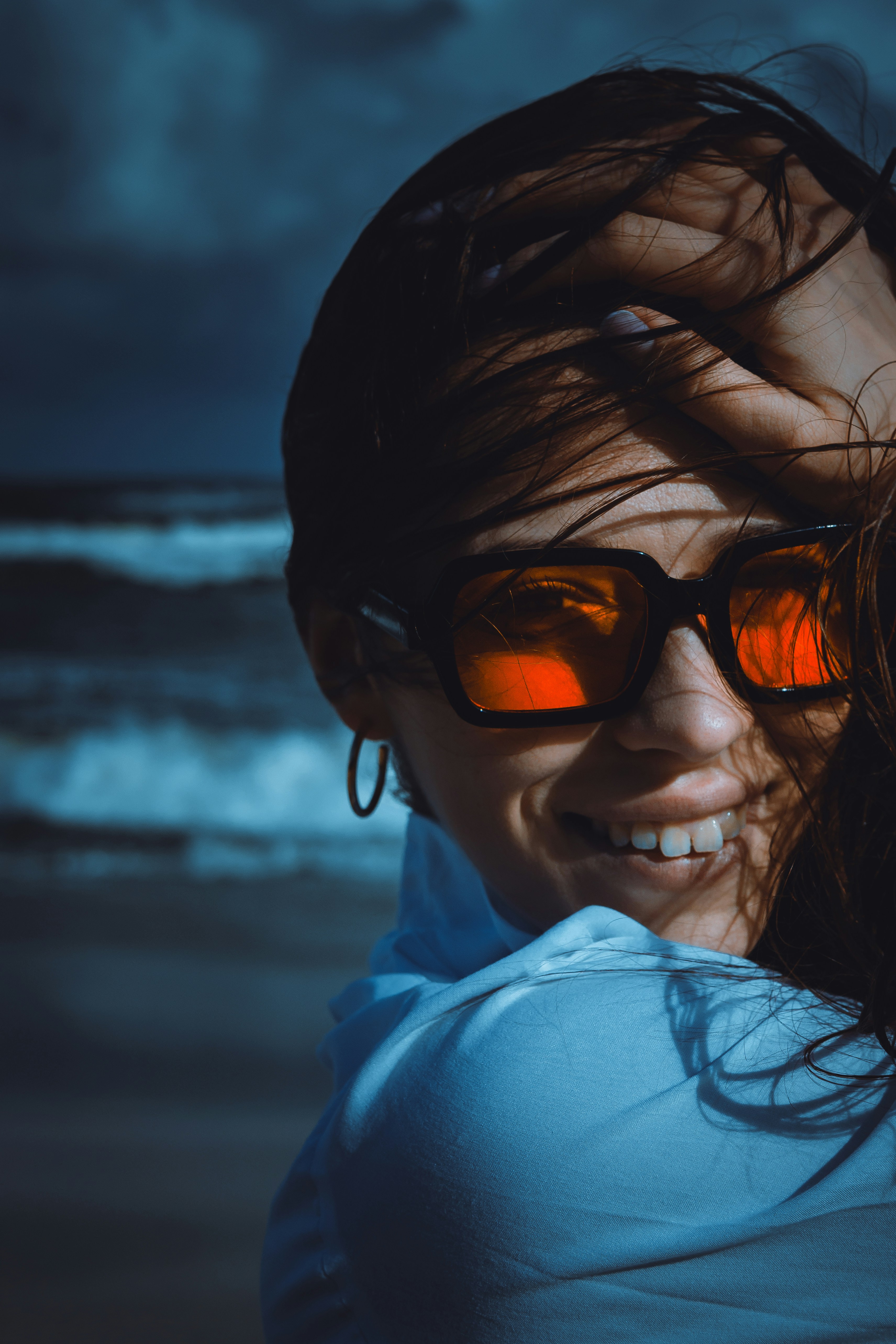 A close-up portrait of a smiling woman wearing reflective orange sunglasses, wind-blown hair, and a light blue jacket at the beach, with dark moody tones in the background.