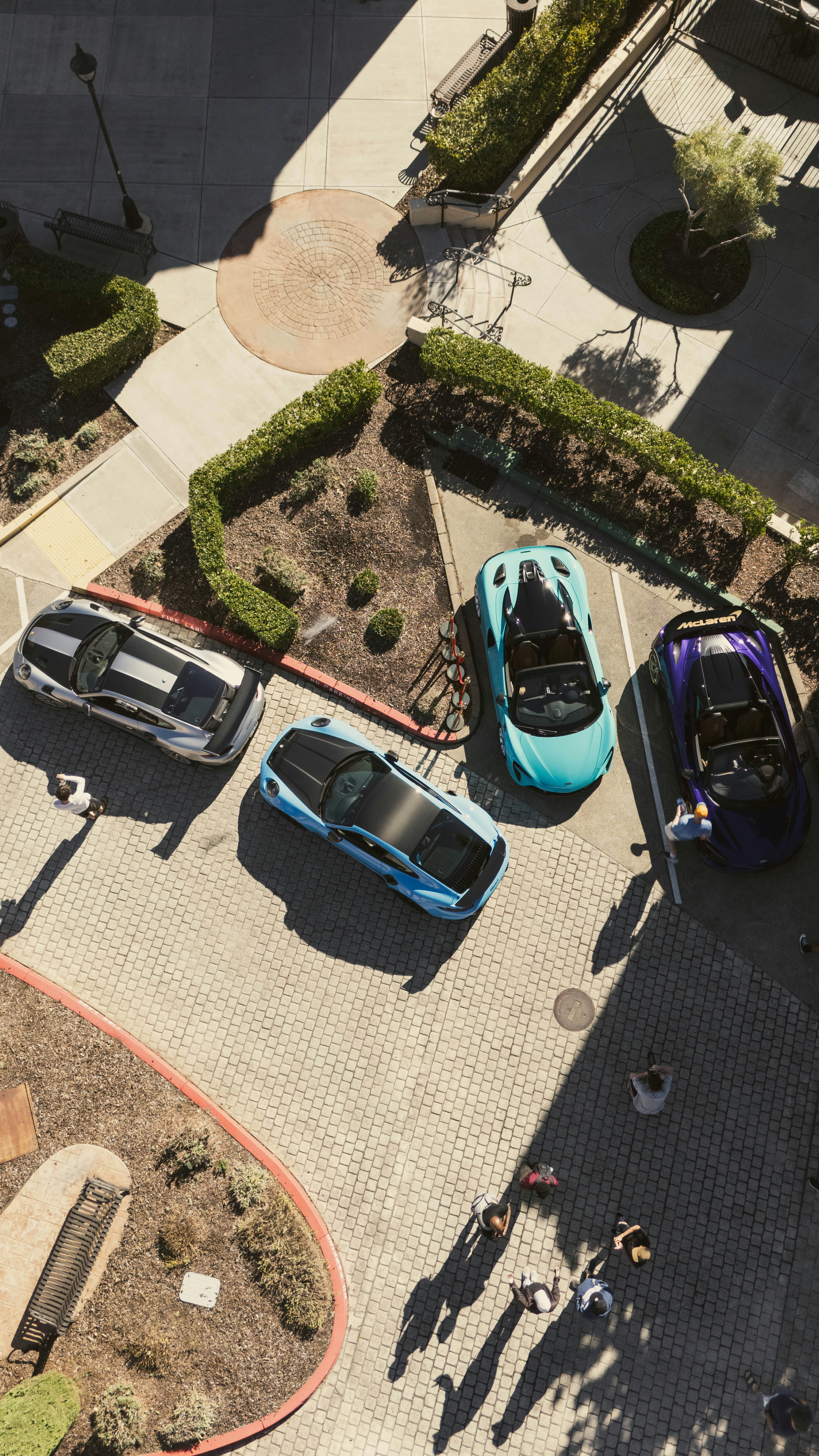 Aerial view of a parking area with colorful sports cars lined up along a curved brick plaza, surrounding hedges and a small group of people casting long shadows on cobblestone pavement.