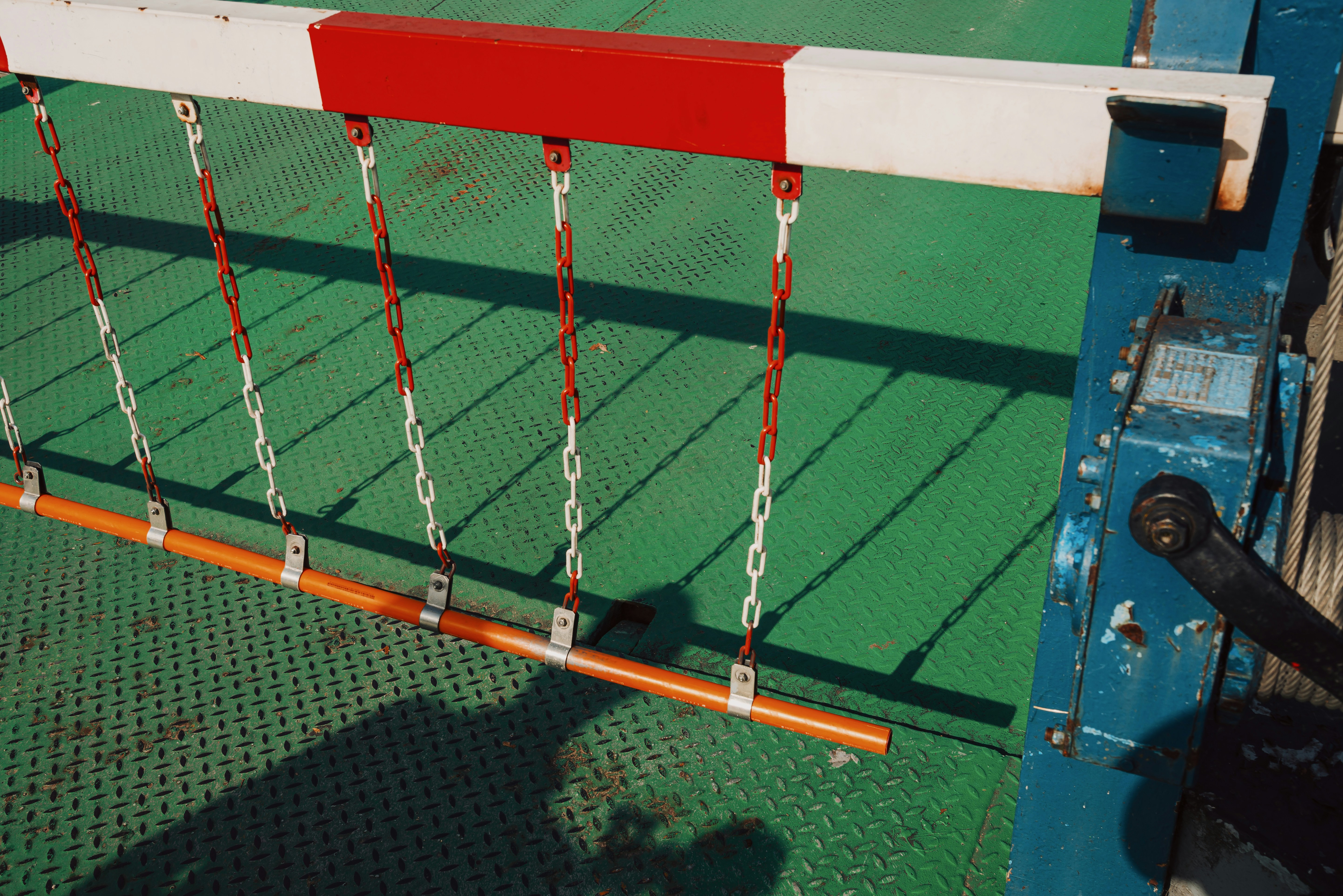 Shadow of a photographer on a green industrial deck with a red-and-white barrier and hanging chains, suggesting a controlled entry area or loading zone. The scene emphasizes texture, color contrast, and industrial geometry.