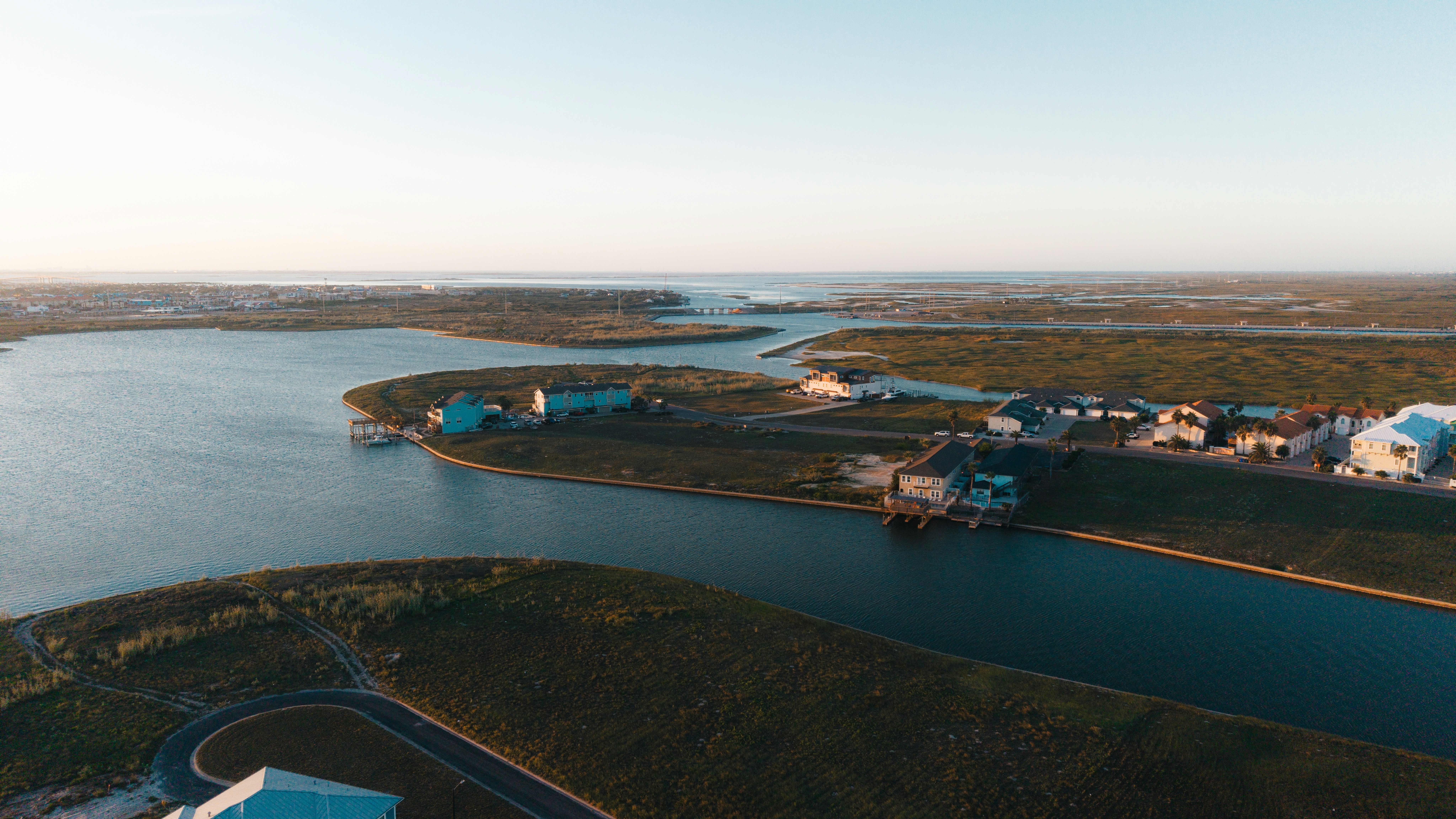 A wide aerial perspective captures the winding waterways and marshland channels weaving through a quiet coastal community. Waterfront houses and private docks line the edges of the water under an expansive sky, highlighting the peaceful balance between residential development and natural coastal terrain.