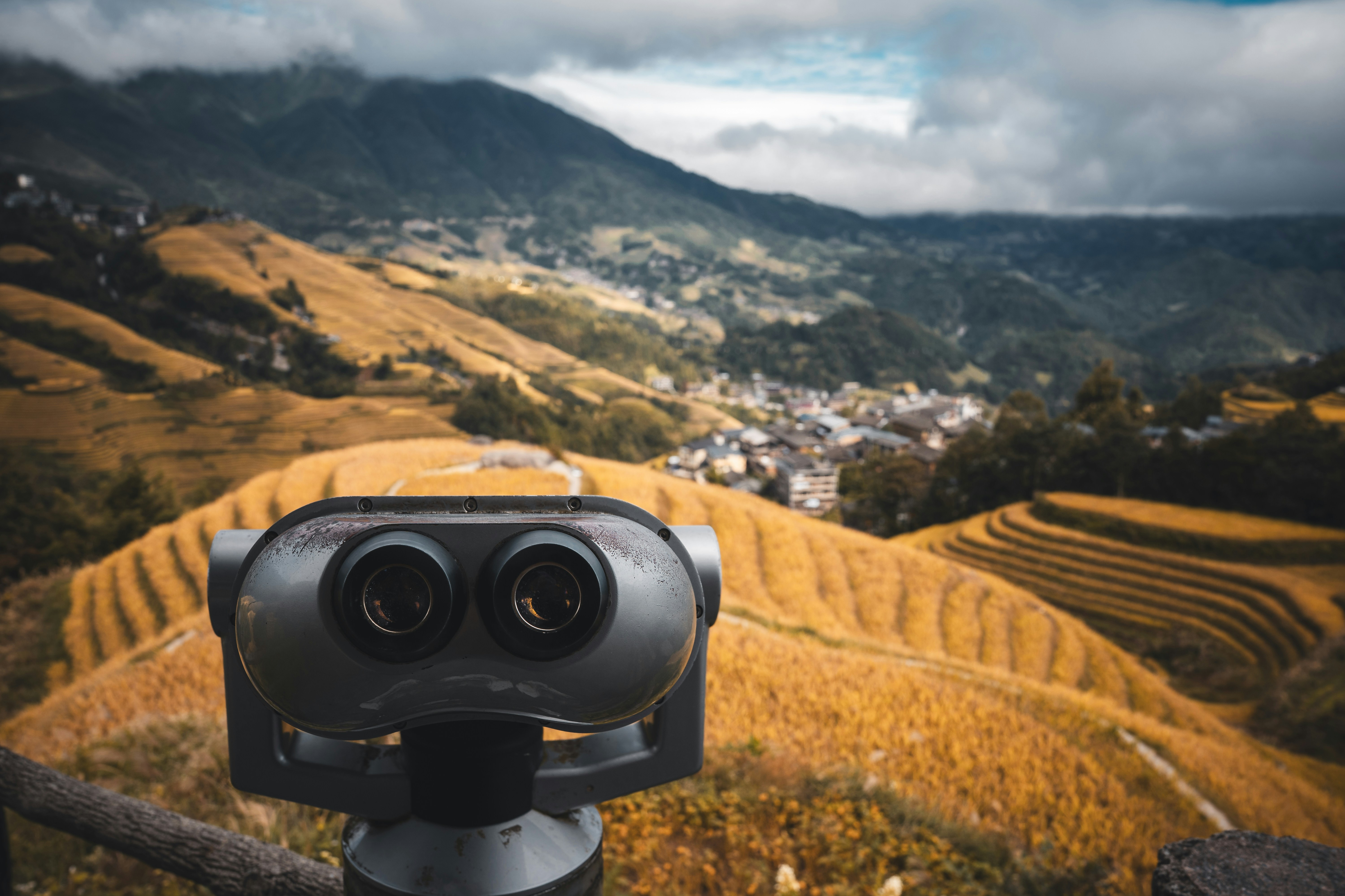 Binoculars mounted in a scenic terraced valley overlook a village with golden fields and distant mountains.